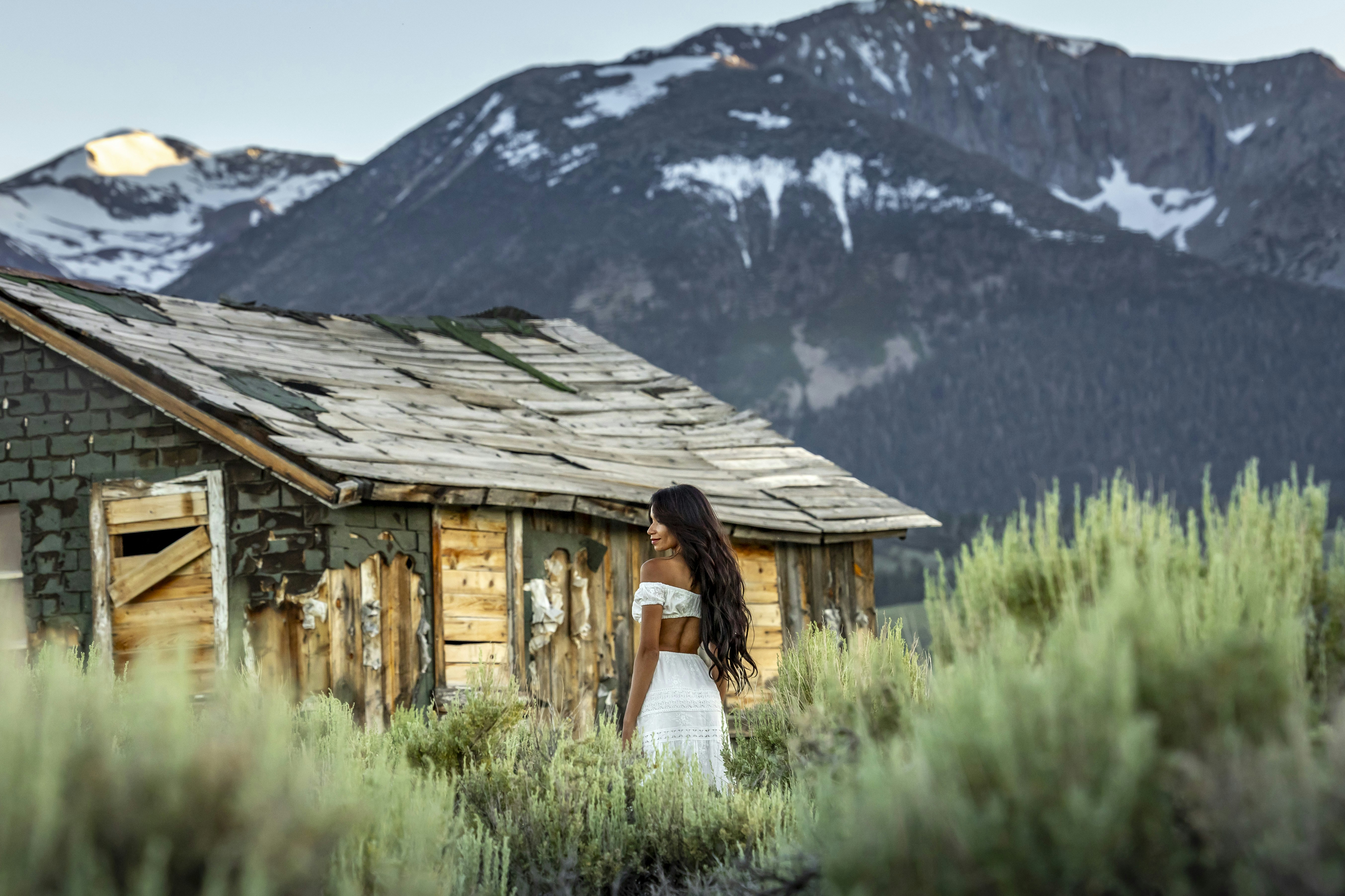 Woman in a white off-shoulder dress stands beside an old wooden chalet in a mountain valley, with a snow-capped range in the background; the scene emphasizes solitude and rustic contrast.