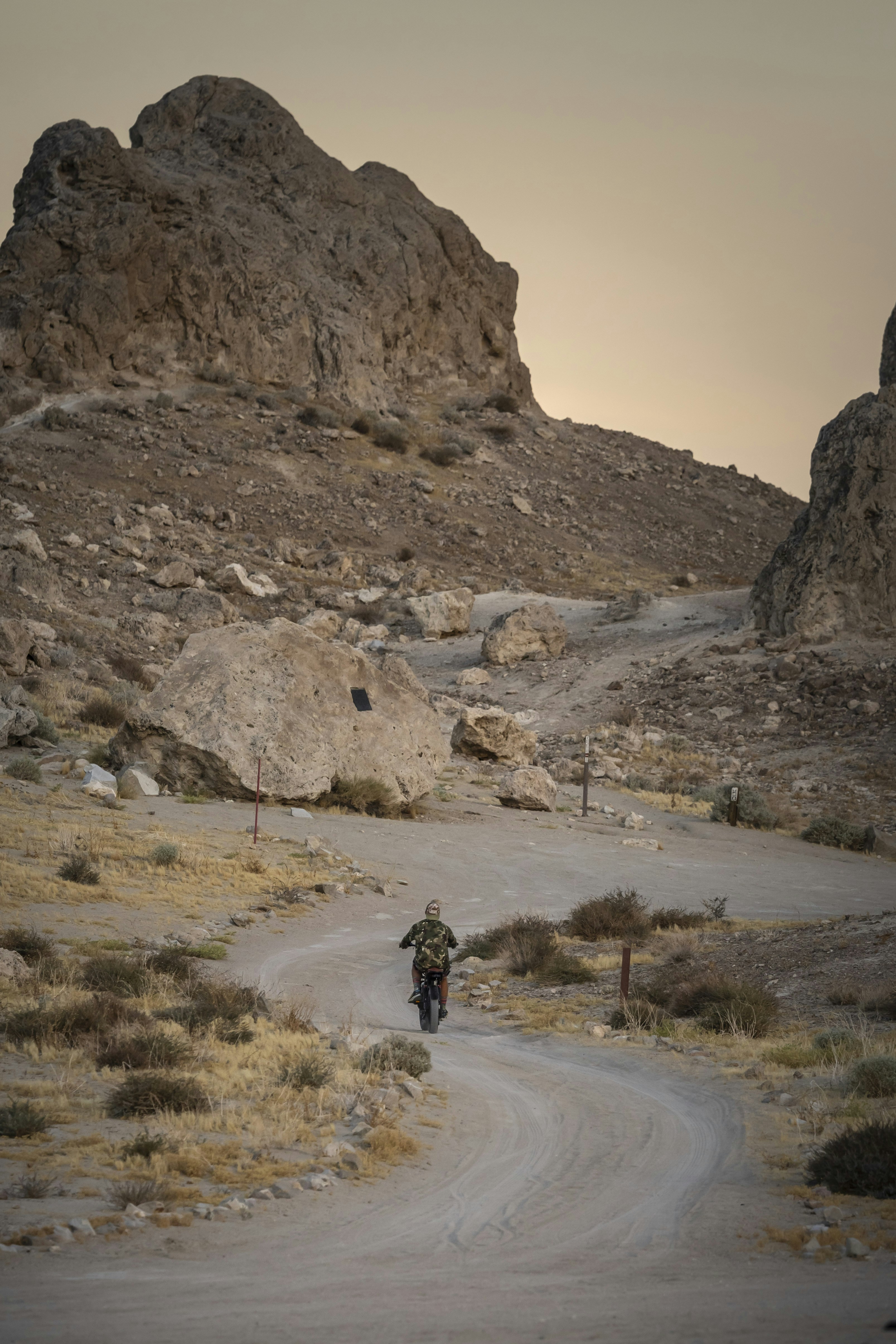 A lone rider navigates a winding dirt road through a rocky desert canyon with warm evening light illuminating rugged cliffs. The scene emphasizes solitude and journey along a remote path.