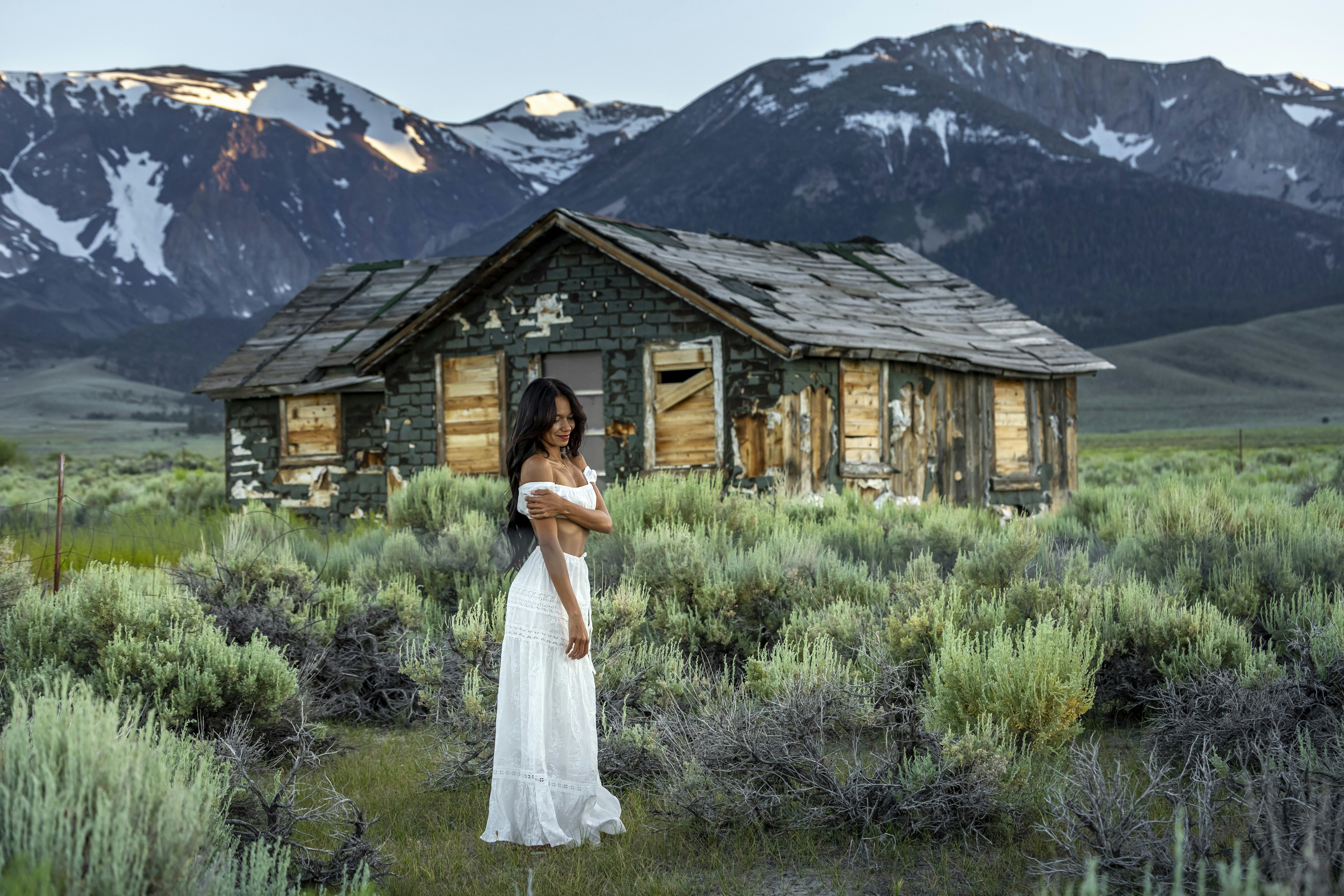 Bride in a white dress stands among sagebrush in front of a weathered cabin with snow-capped mountains behind.