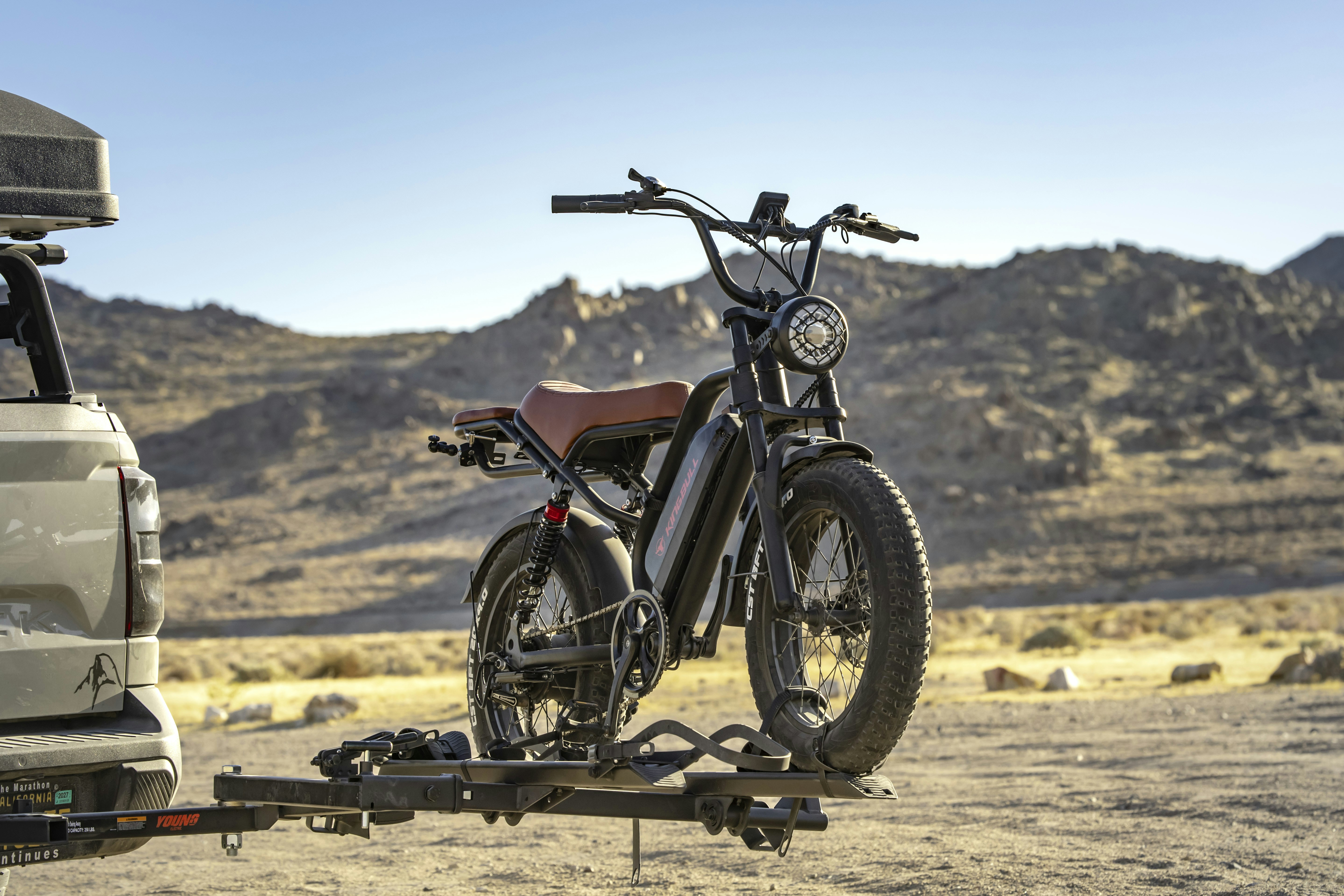 Electric motorcycle on a flatbed trailer in a desert landscape with rugged hills in the background; a compact bike with brown seat and black frame. A logo or badge may be present on the bike; scene resembles a product/vehicle setup rather than a staged portrait.
