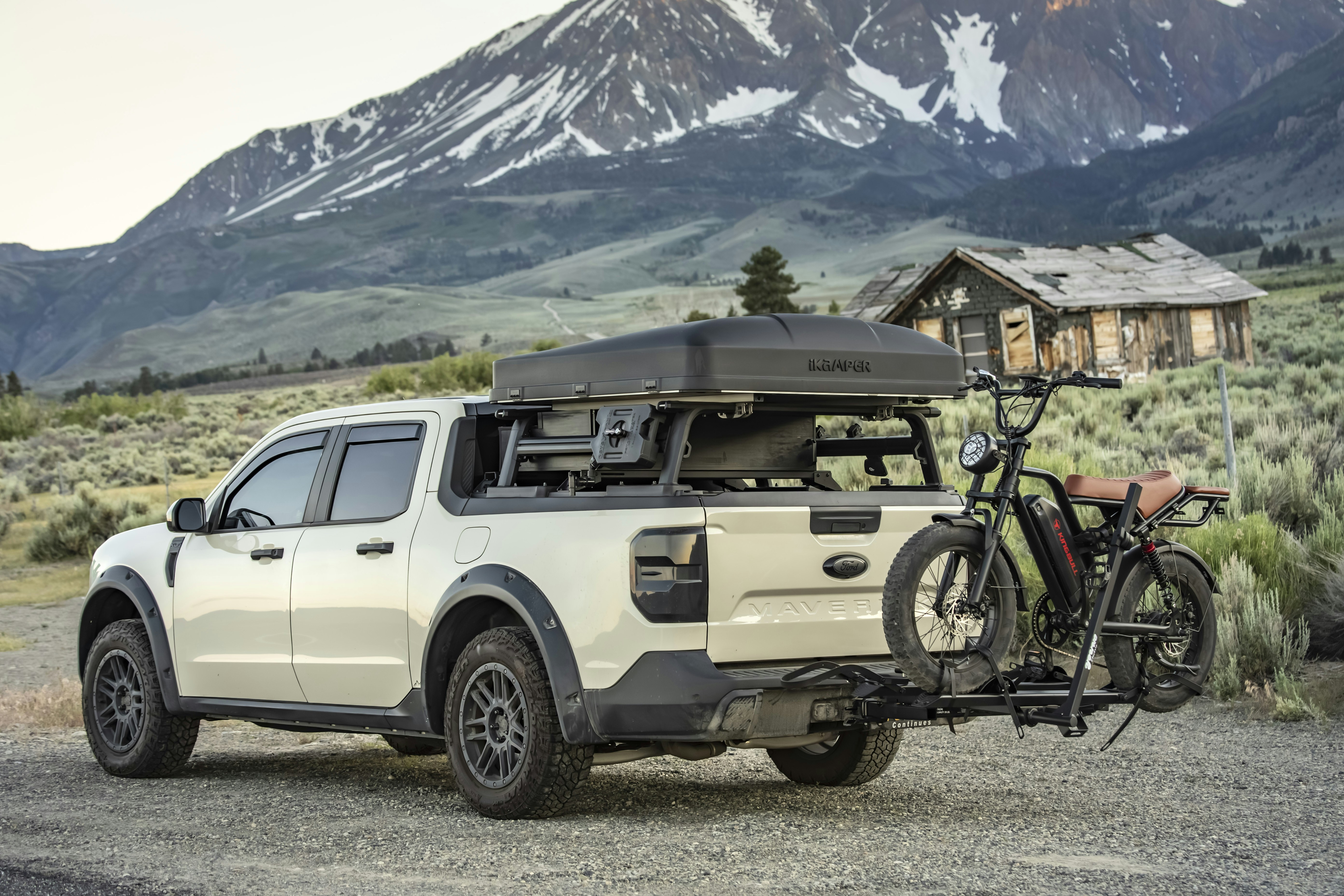 White pickup with rooftop cargo and mounted bike sits on a gravel lot beside a rustic cabin, with a snow-dusted mountain range in the background.