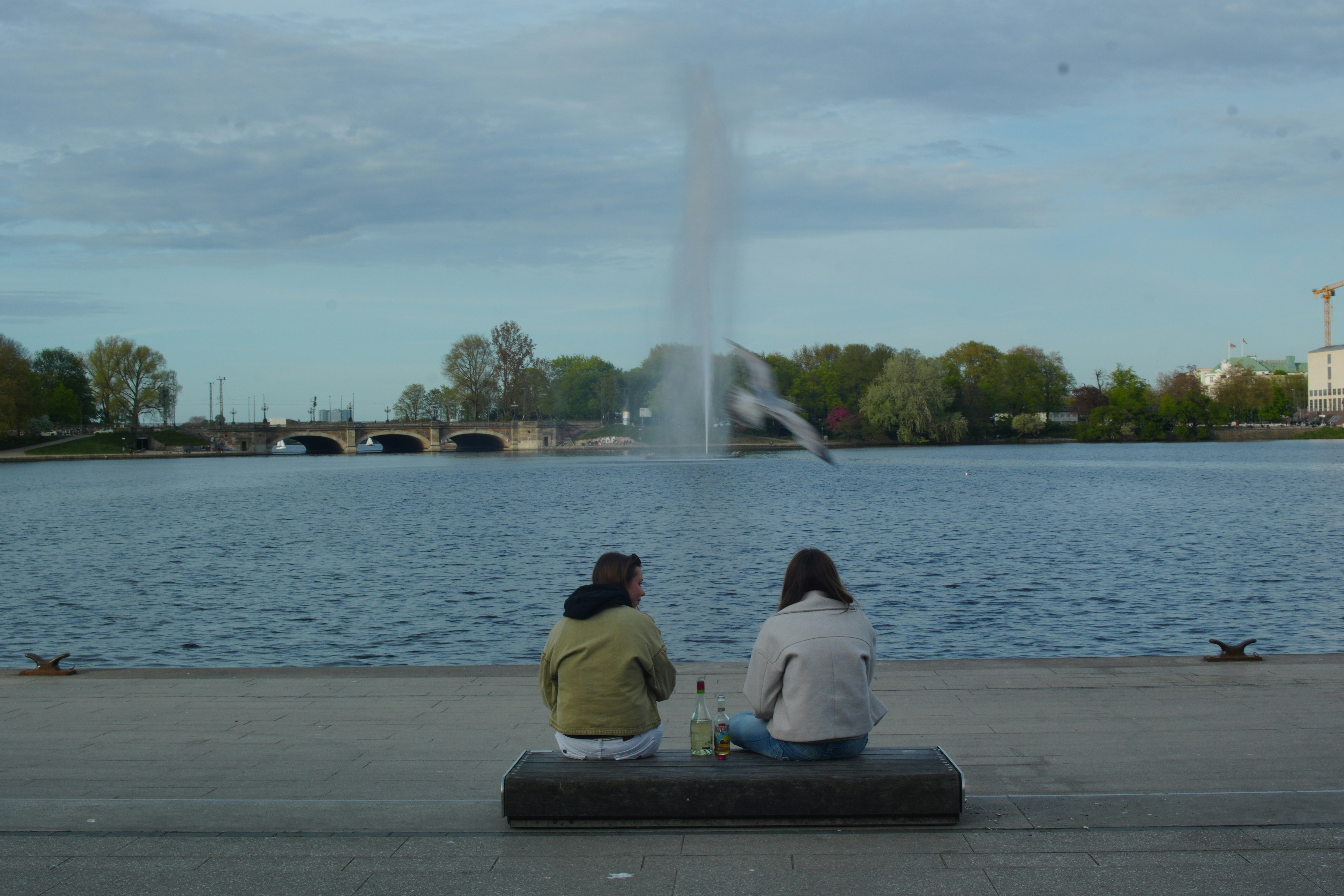 Pause by the Lake: Two Friends Share a Quiet Moment with a Backdrop of a Dancing Fountain