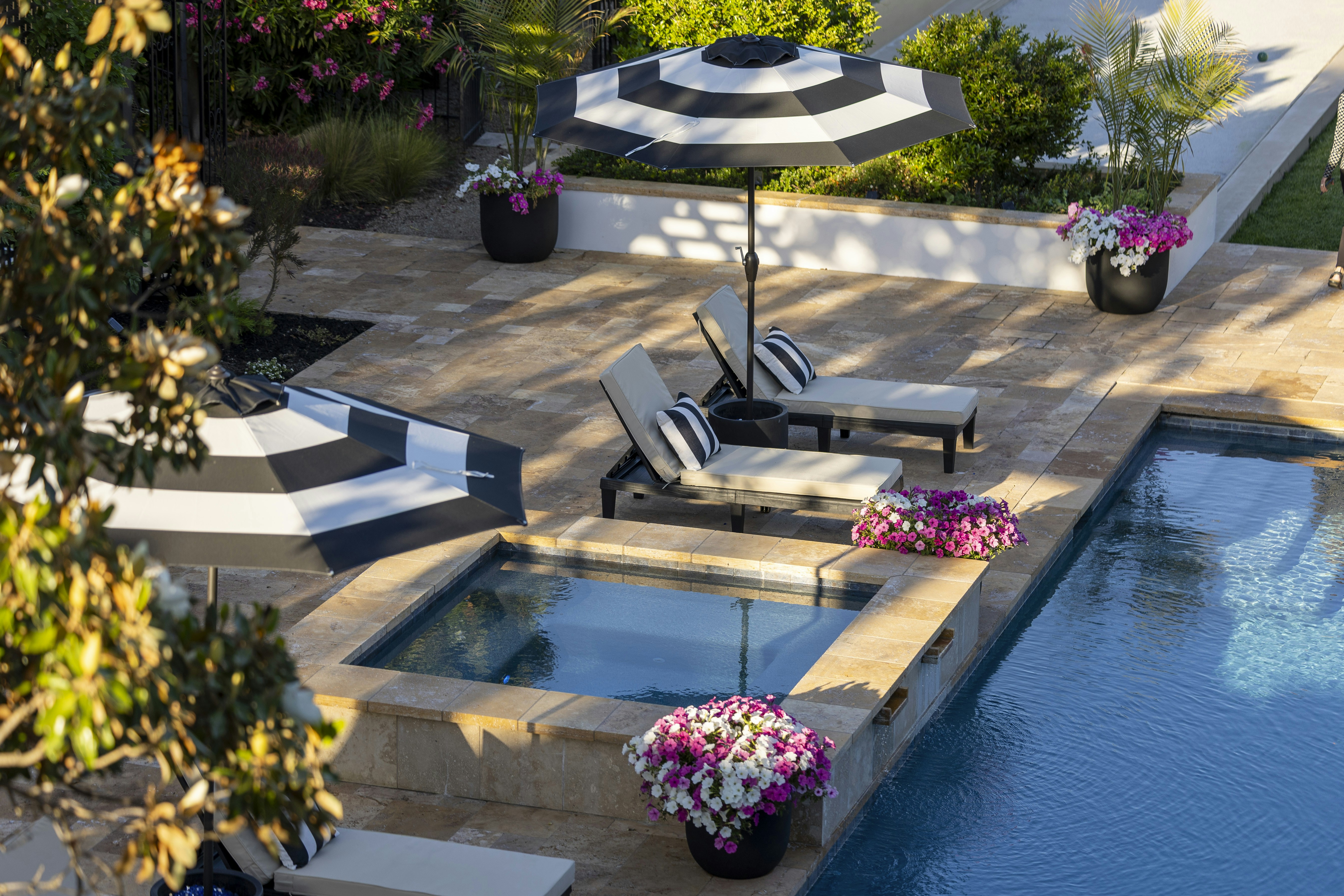 Sunlit poolside terrace with striped umbrellas, lounge chairs, and potted flowers along a beige stone deck.