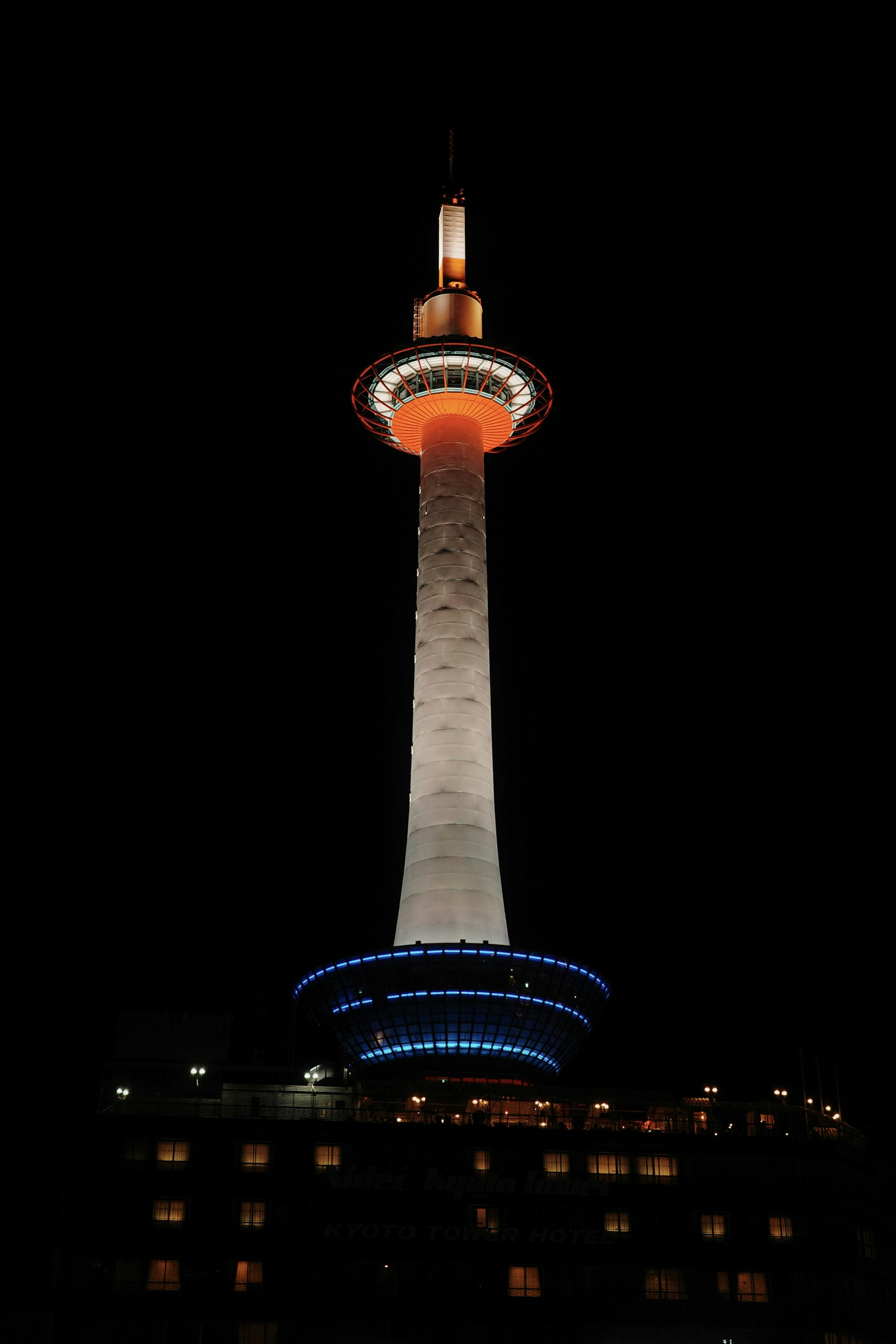 Brightly lit broadcast tower rises from a dark cityscape, rings of orange and blue glow encircling the midsection. The photo emphasizes vertical structure against a black sky and a hint of illuminated windows below.