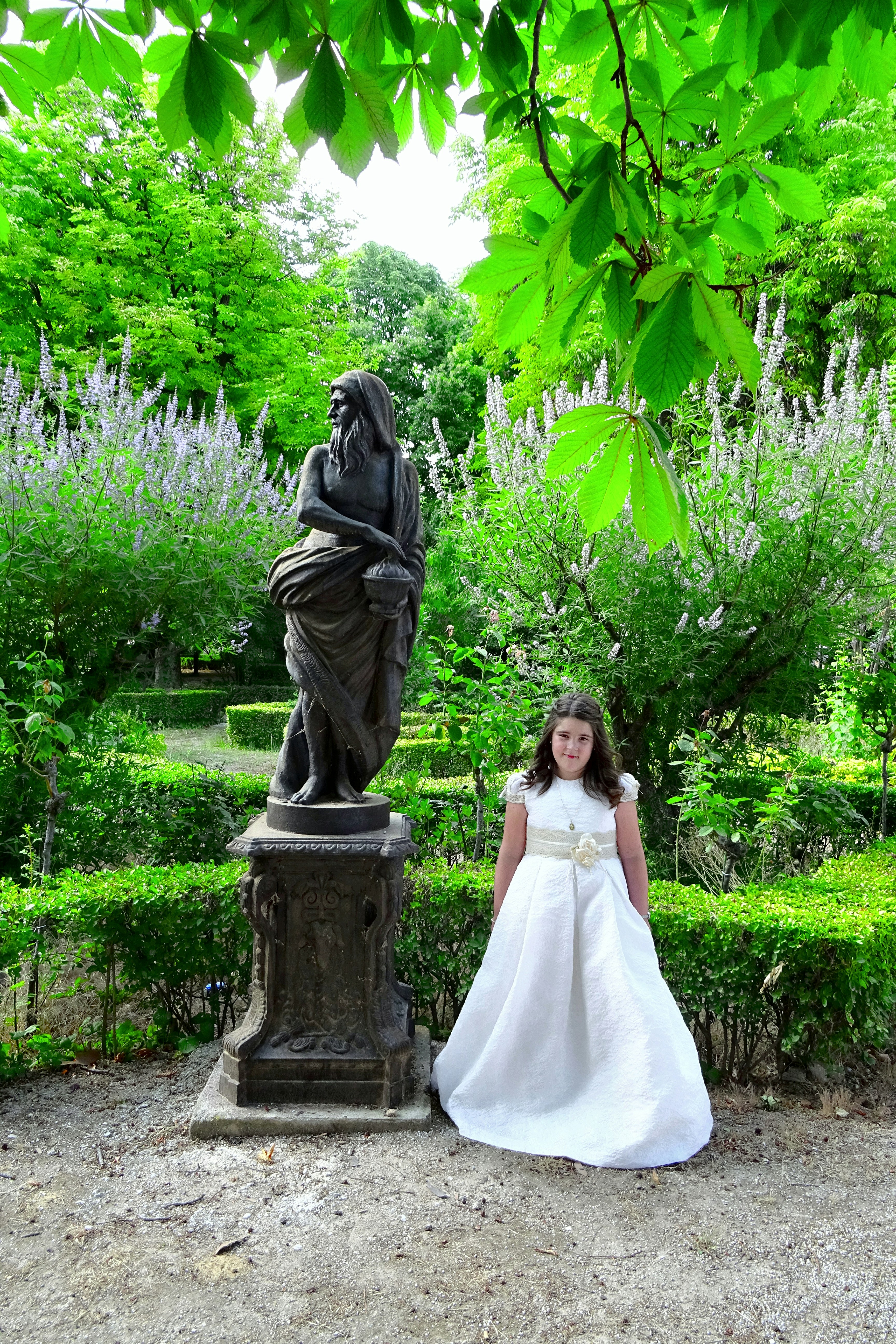 Girl in a White Dress beside a Statue in a Lush Park