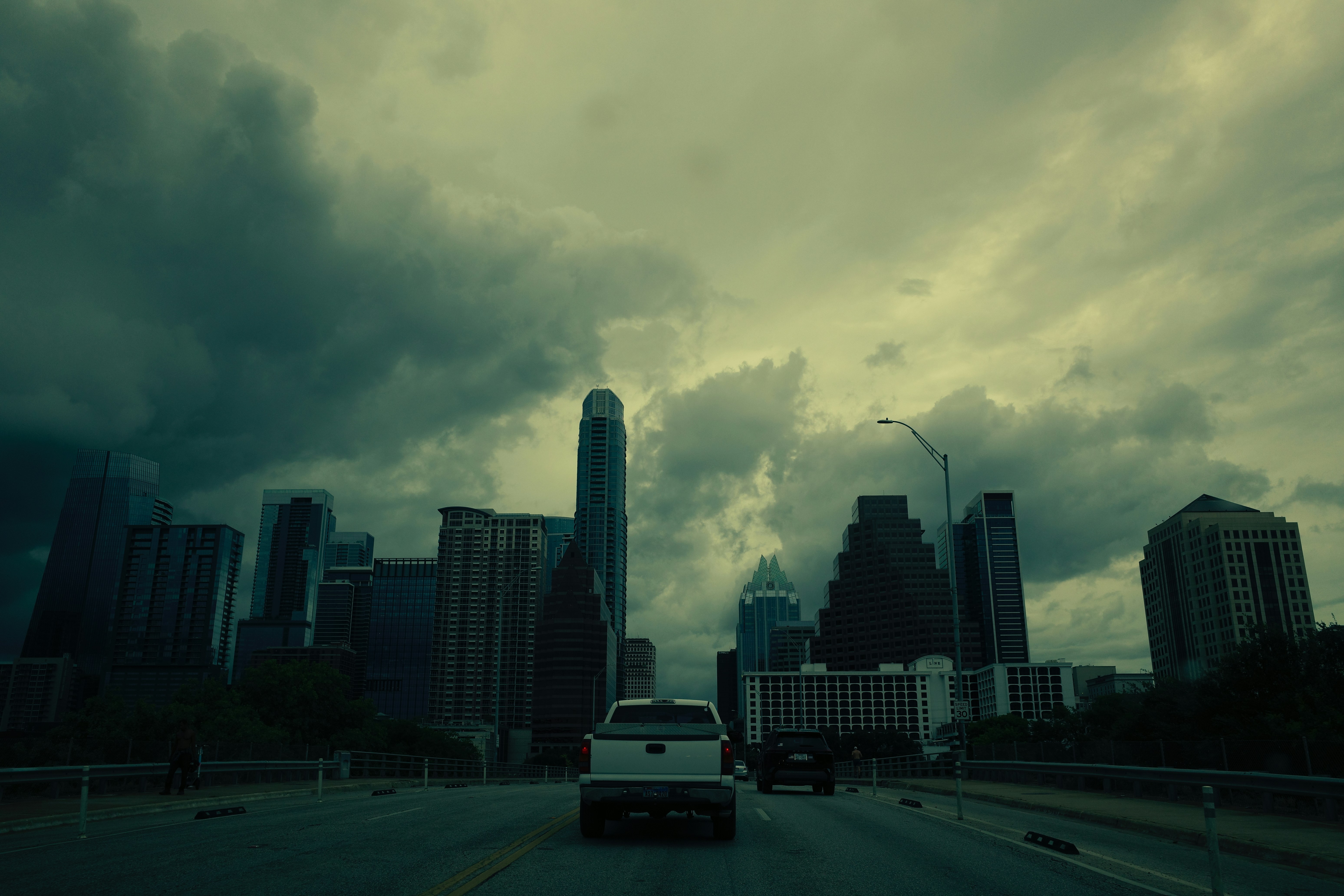 Steel skyline under heavy clouds seen from a highway with vehicles in the foreground; a moody urban scene emphasizing perspective and weather.