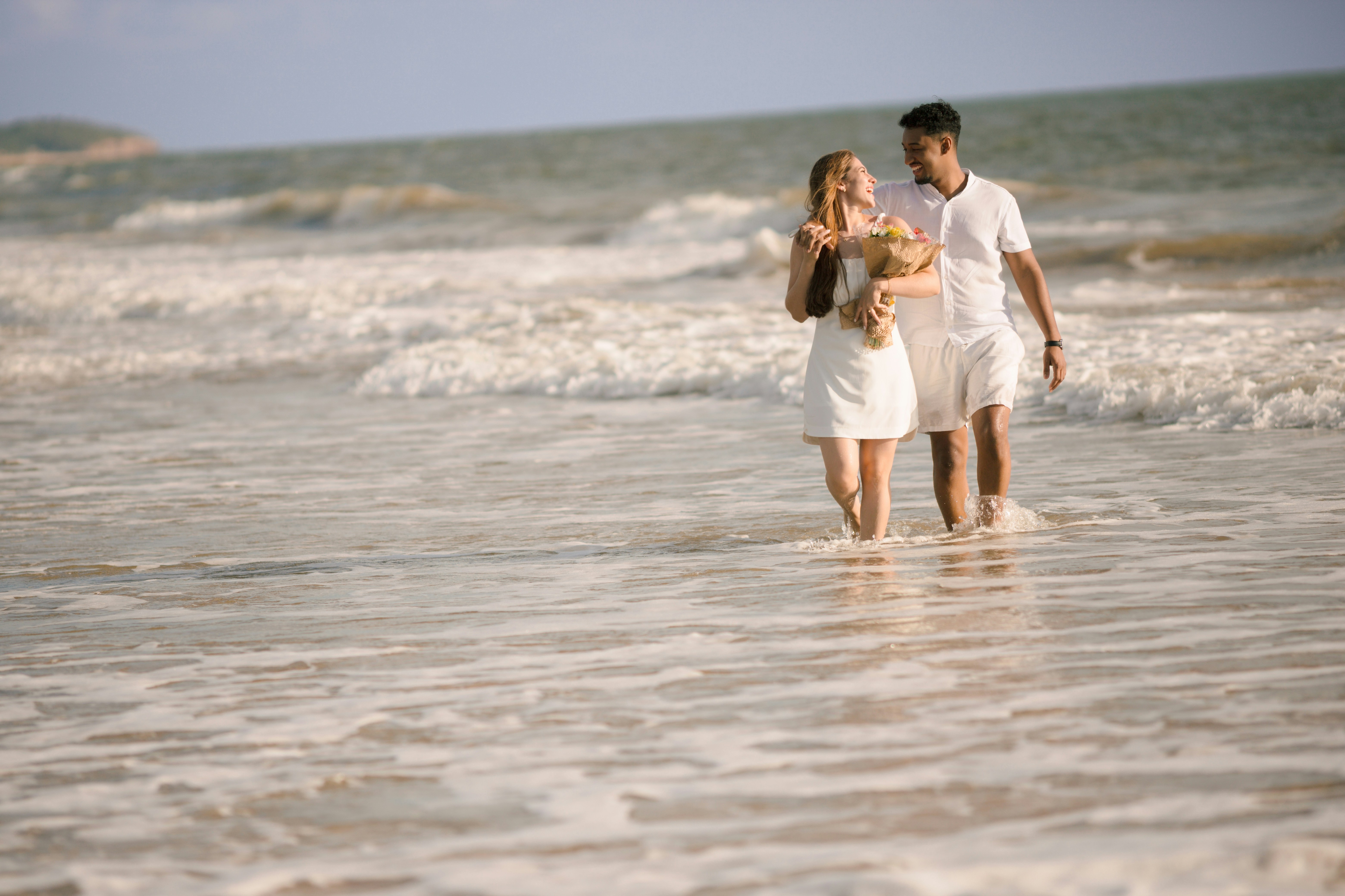 Beachside Bouquet: Two Lovers in White Walk Along the Tidal Edge