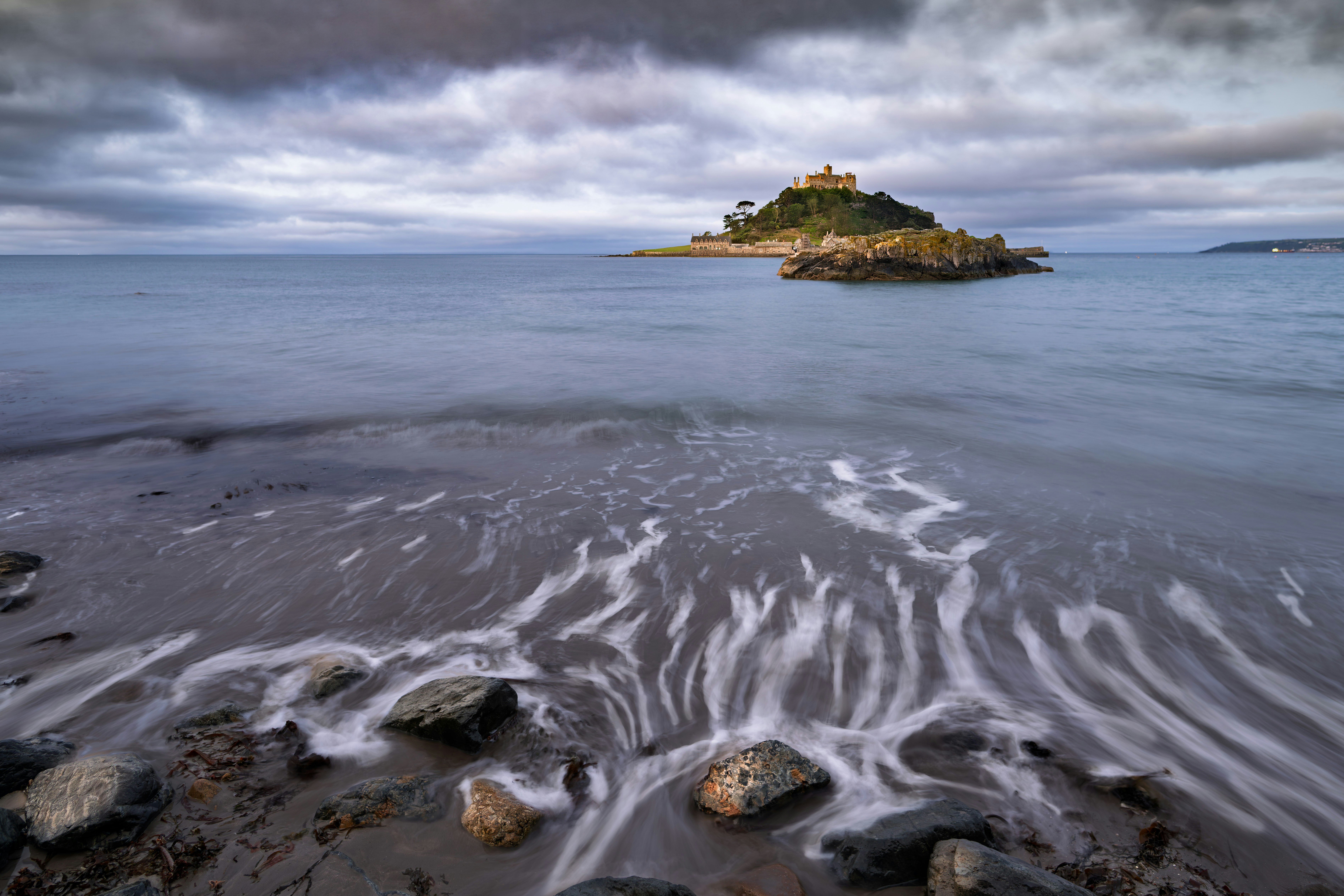 A moody pre-dawn view of Saint Michael’s Mount rising offshore from Marazion, with storm clouds and soft long-exposure surf creating a dramatic Cornish seascape.