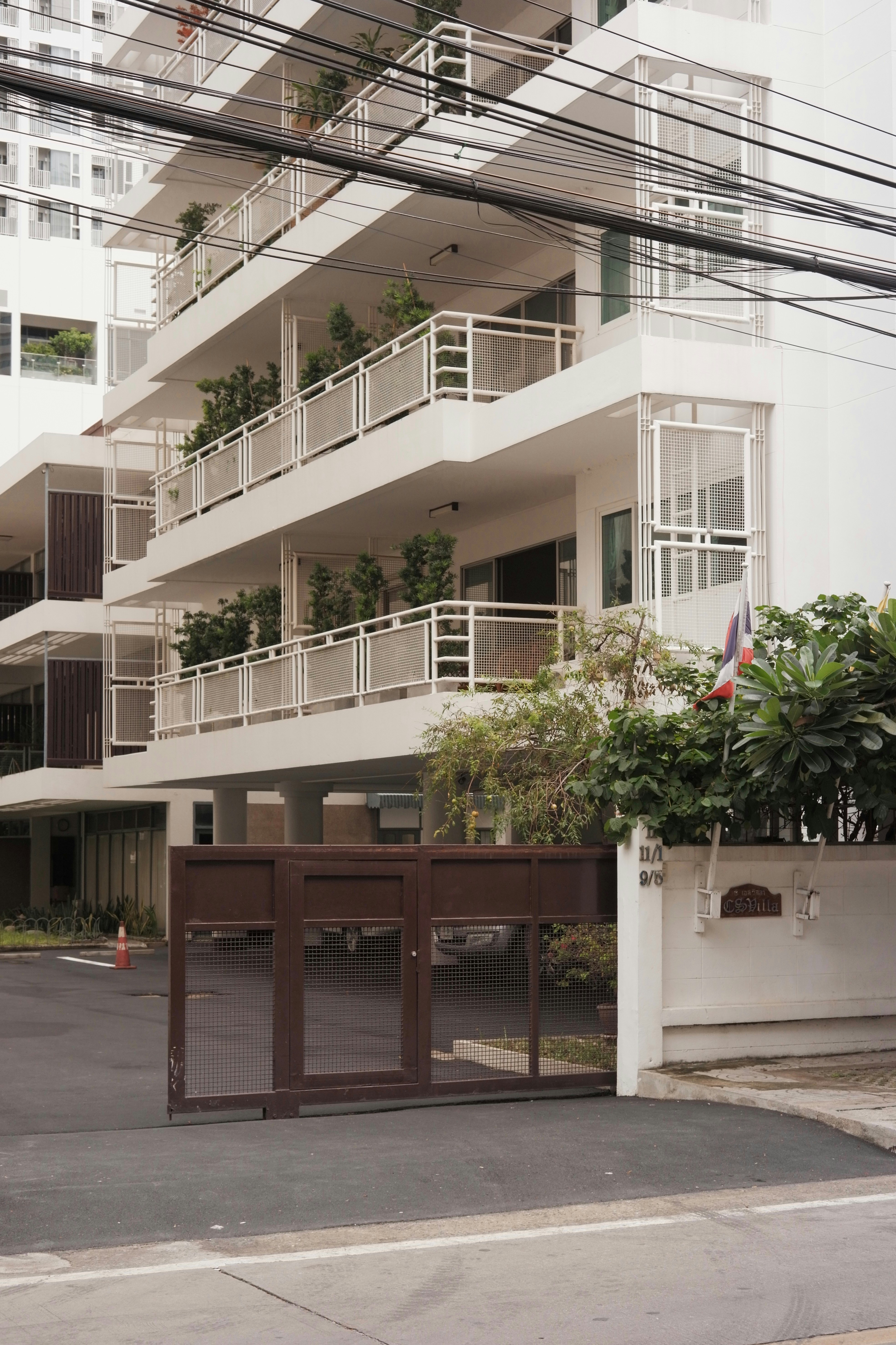 A modern residential building with stepped balconies lined with greenery, gated entry, and a Thai flag near the entrance.