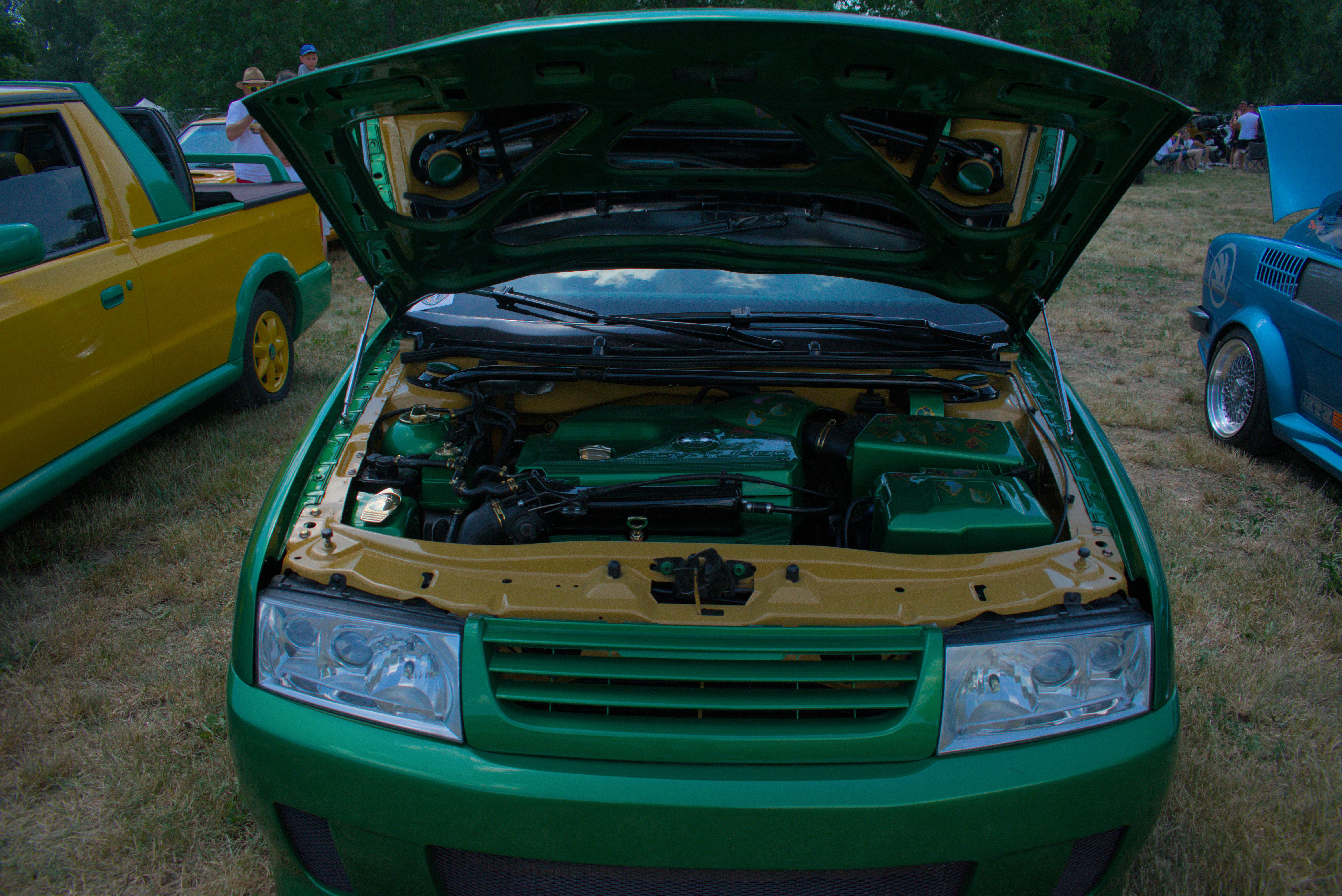 Green car with its hood open at a car show, engine bay visible and other colorful vehicles in the background.