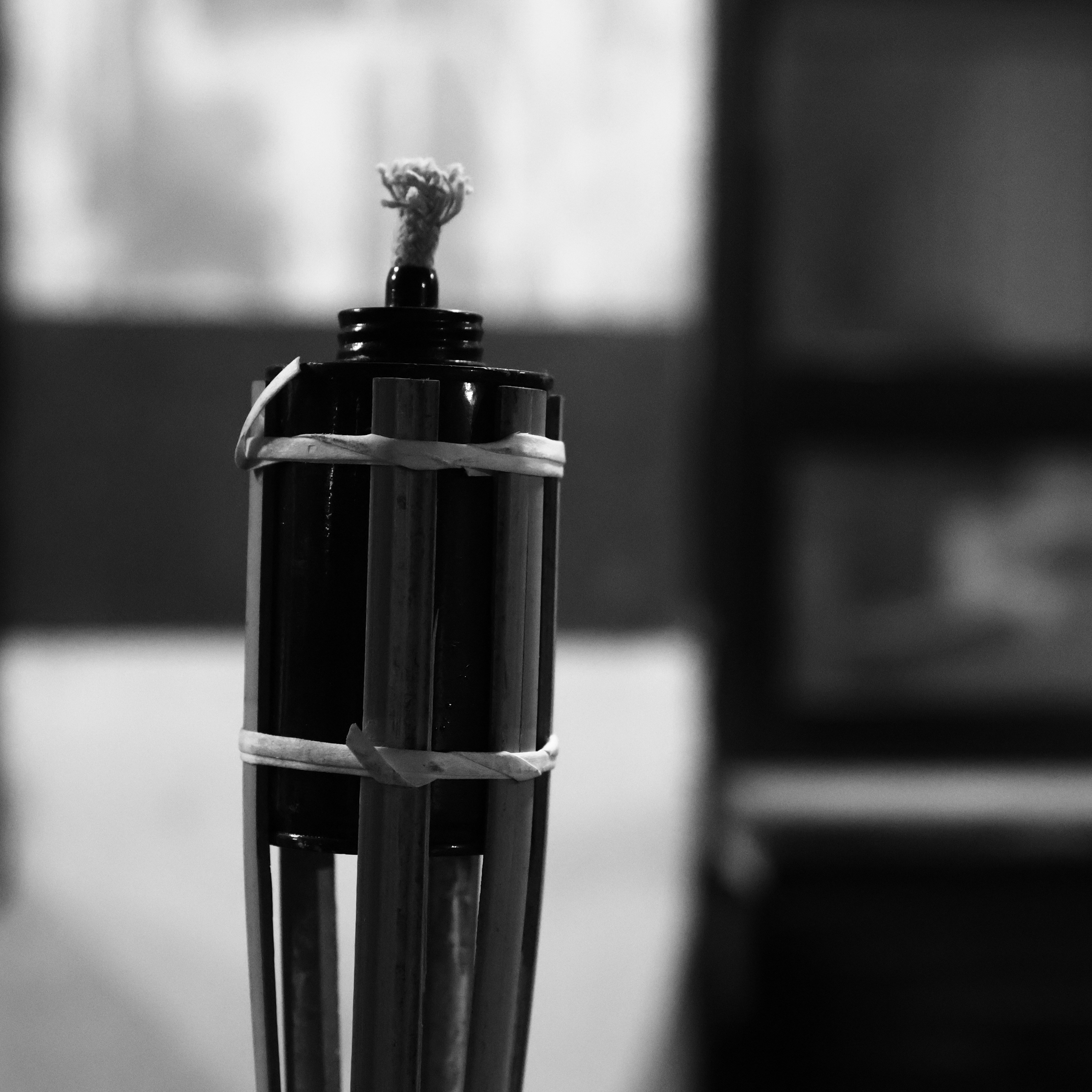 Monochrome still life of a bamboo-wound torch-like object with a charred wick at the top, set against a soft, blurred background. The composition focuses on the central object’s vertical form and texture.