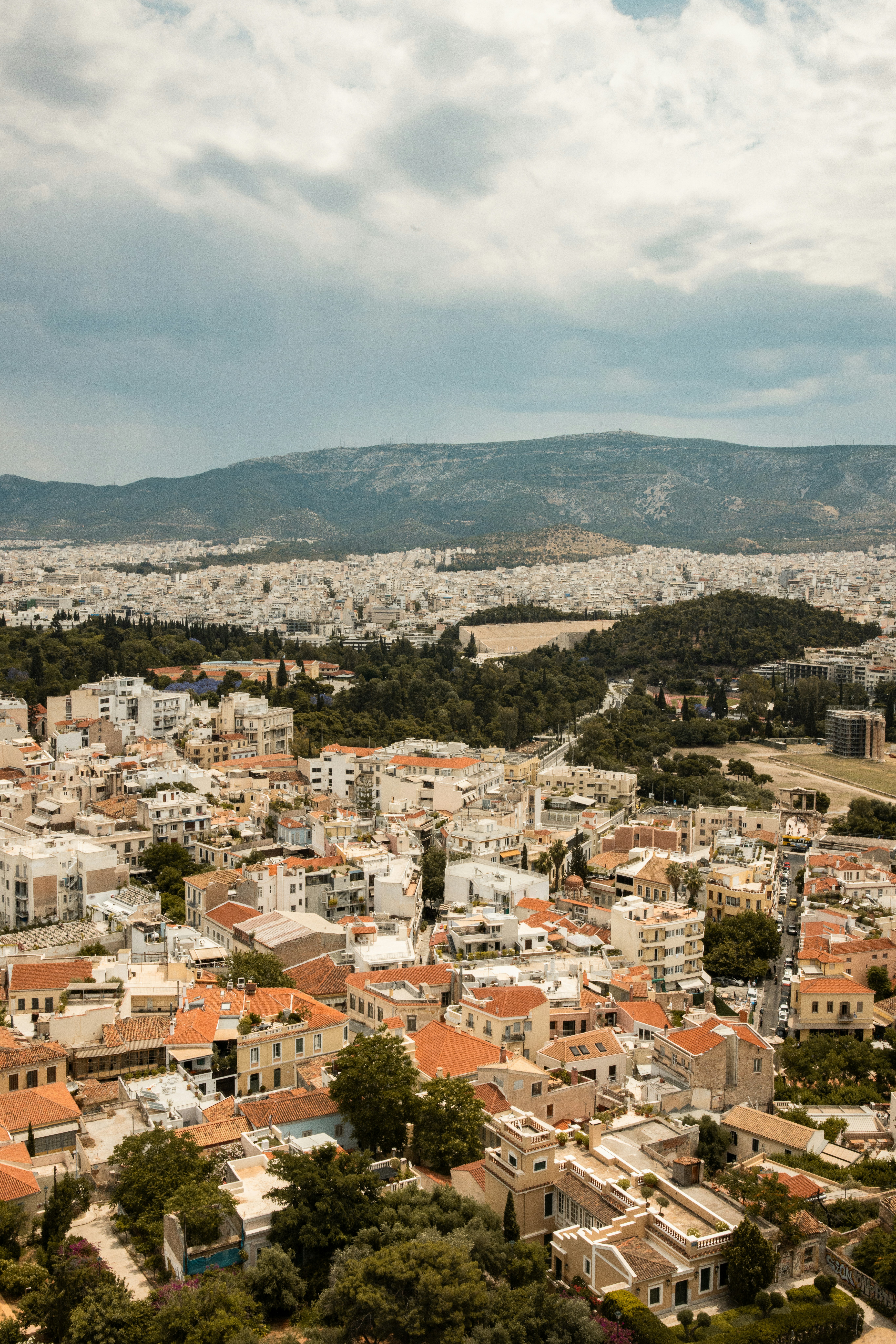 Aerial view of a sunlit Mediterranean town with terracotta roofs cascading toward greener hills, under a cloudy sky. This is a landscape photograph capturing the dense housing and surrounding vegetation.