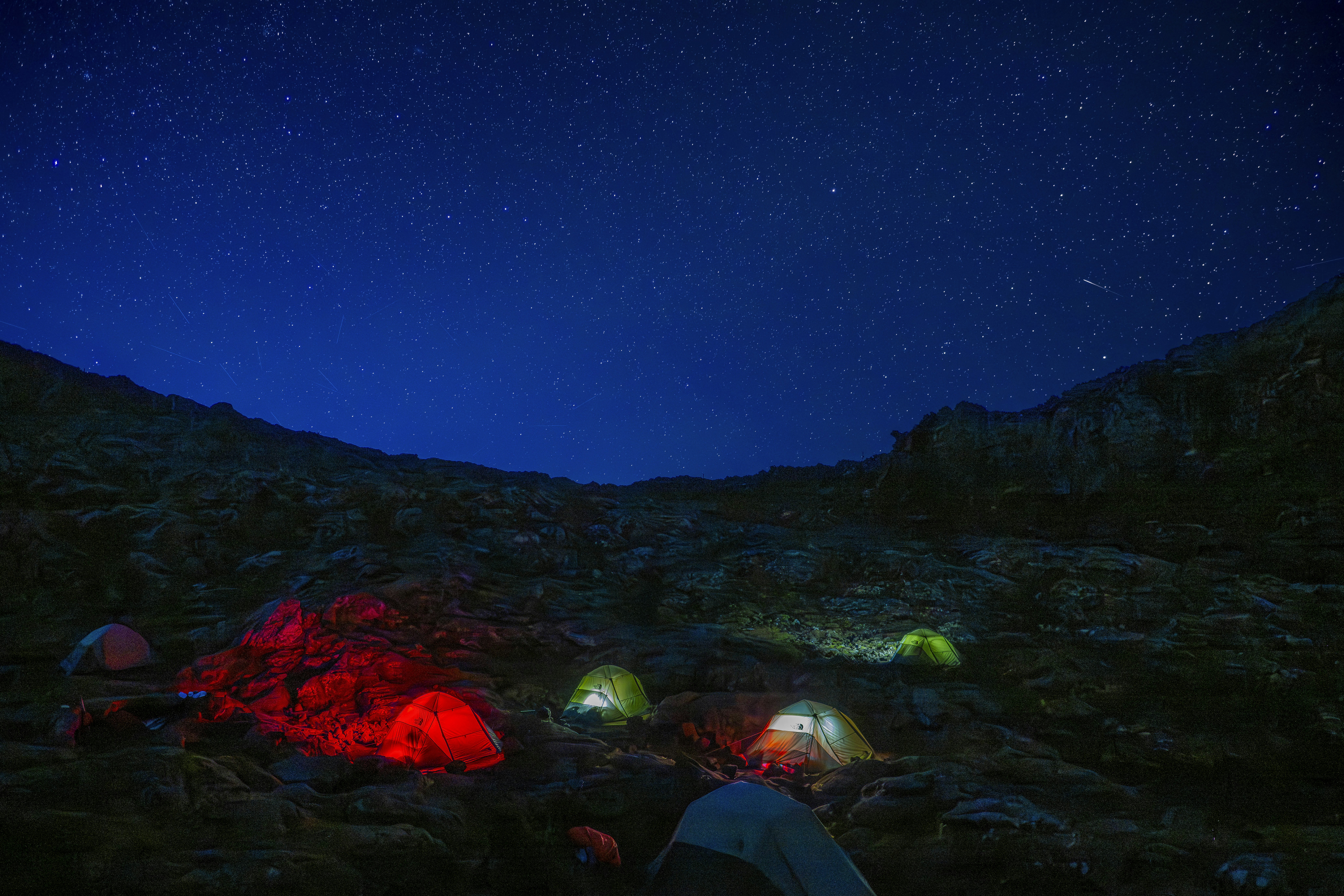 Night-time campsite speckled with red and yellow tents beneath a star-filled sky; rugged terrain and a calm, dark landscape set the scene for stargazing.