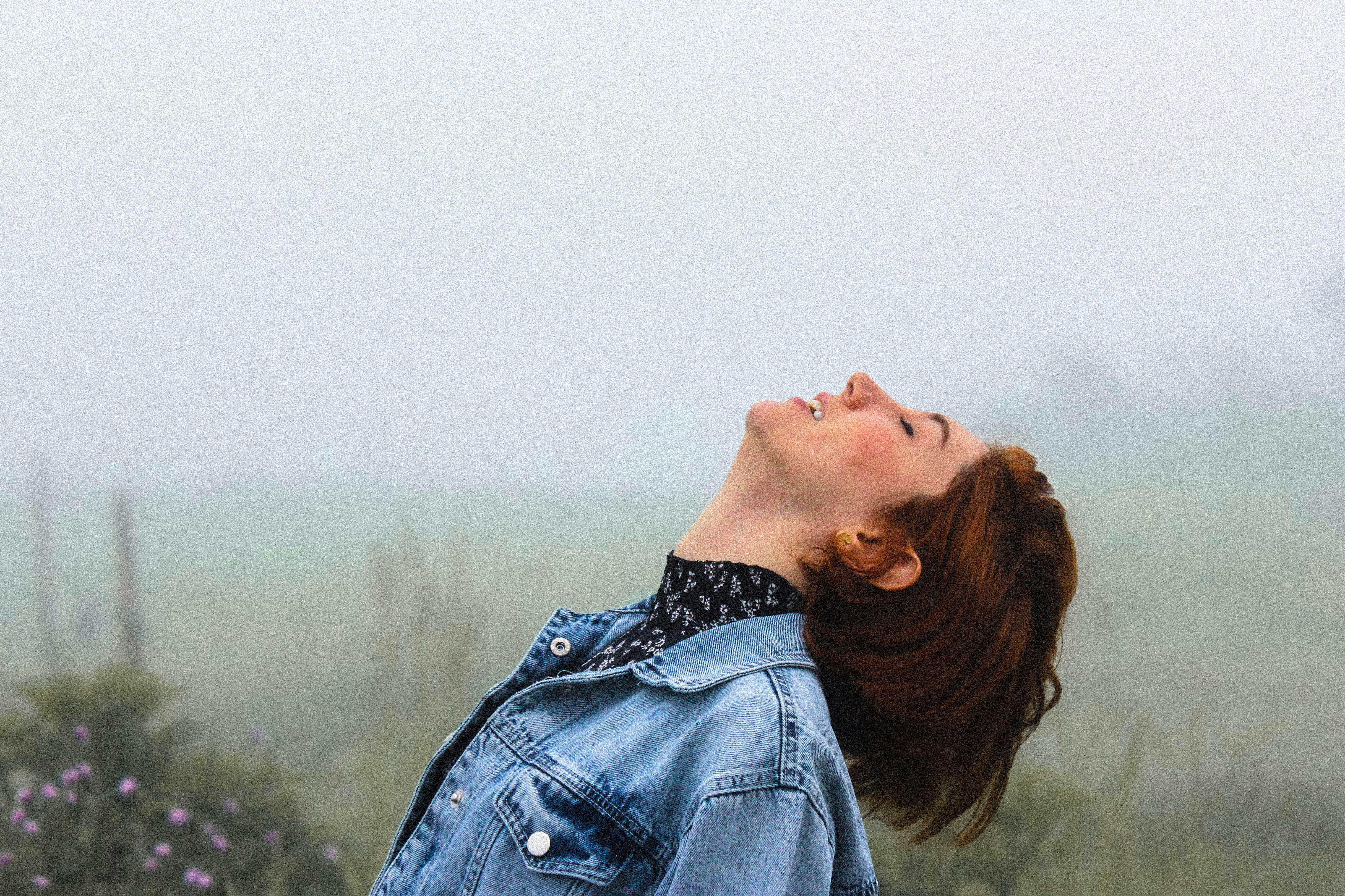 A woman in a denim jacket tilts her head back toward a pale sky, suggesting a moment of release or contemplation in a misty, open field.