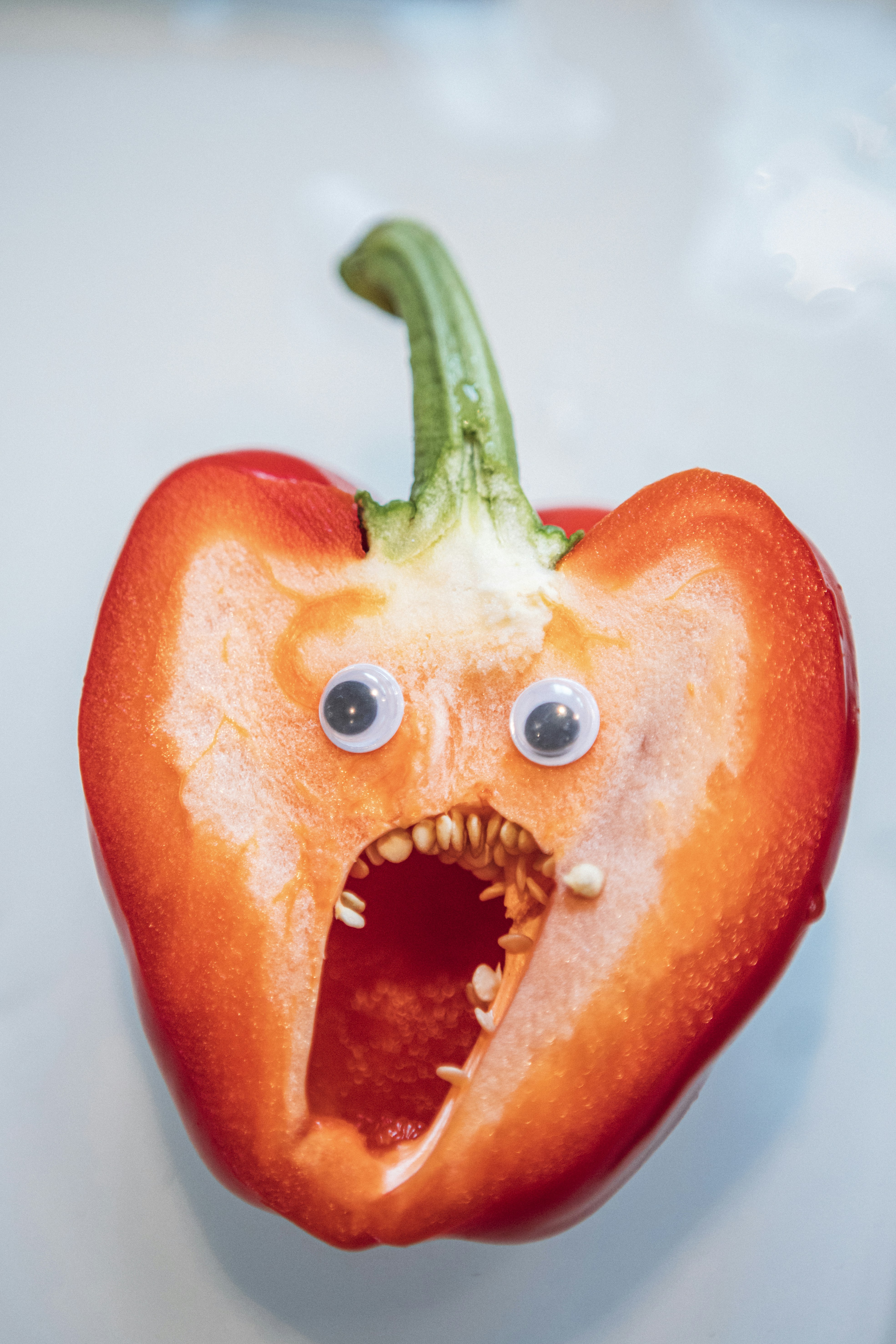 A cute bell pepper carved with eyes and a wide open mouth, giving it a surprised expression; the scene features a single pepper cut in half with googly eyes and a jagged interior, set against a clean background.