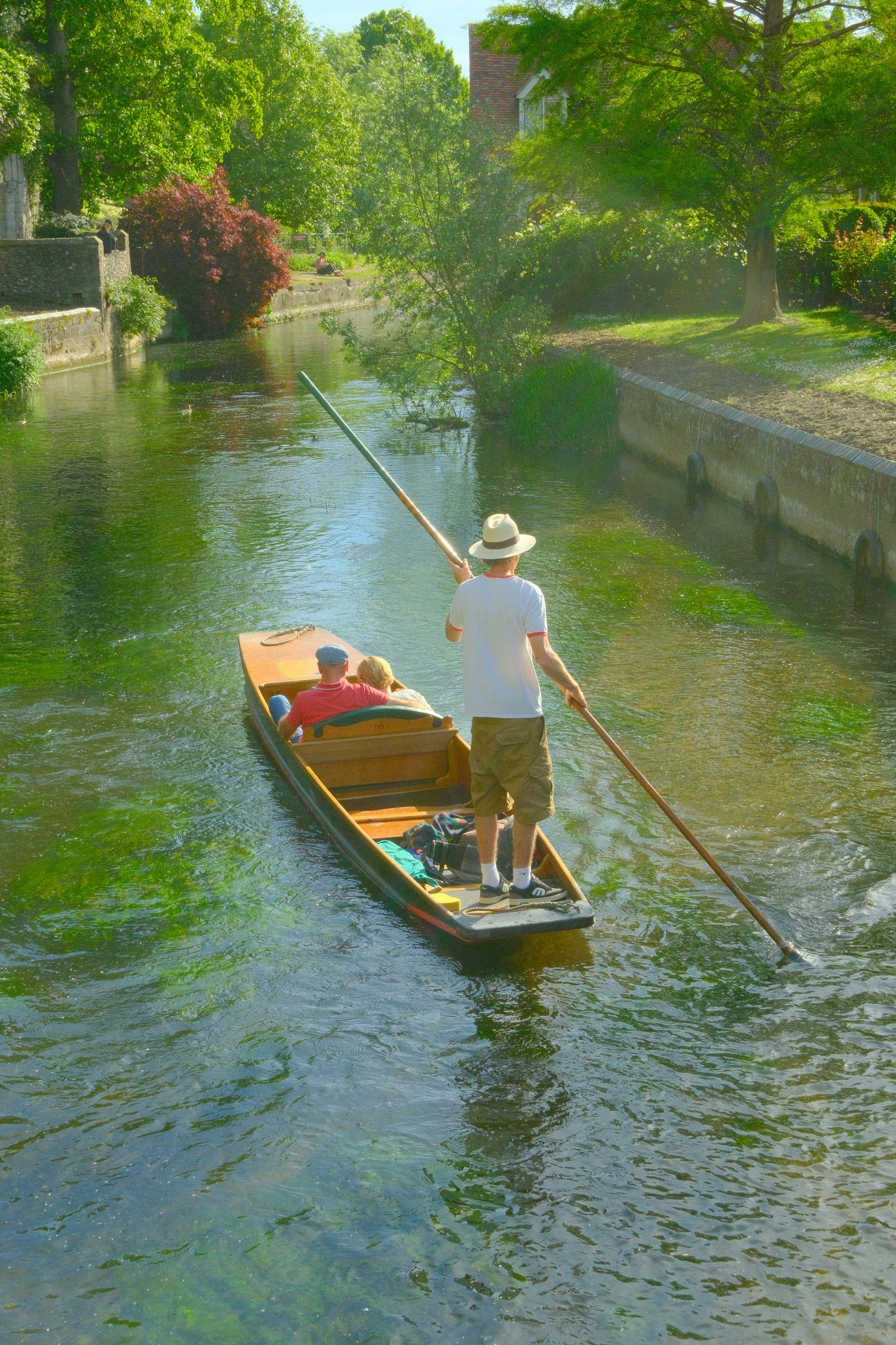 Three people ride a longboat through a sun-dappled canal, man standing at stern with a long pole guiding the craft along verdant embankments.