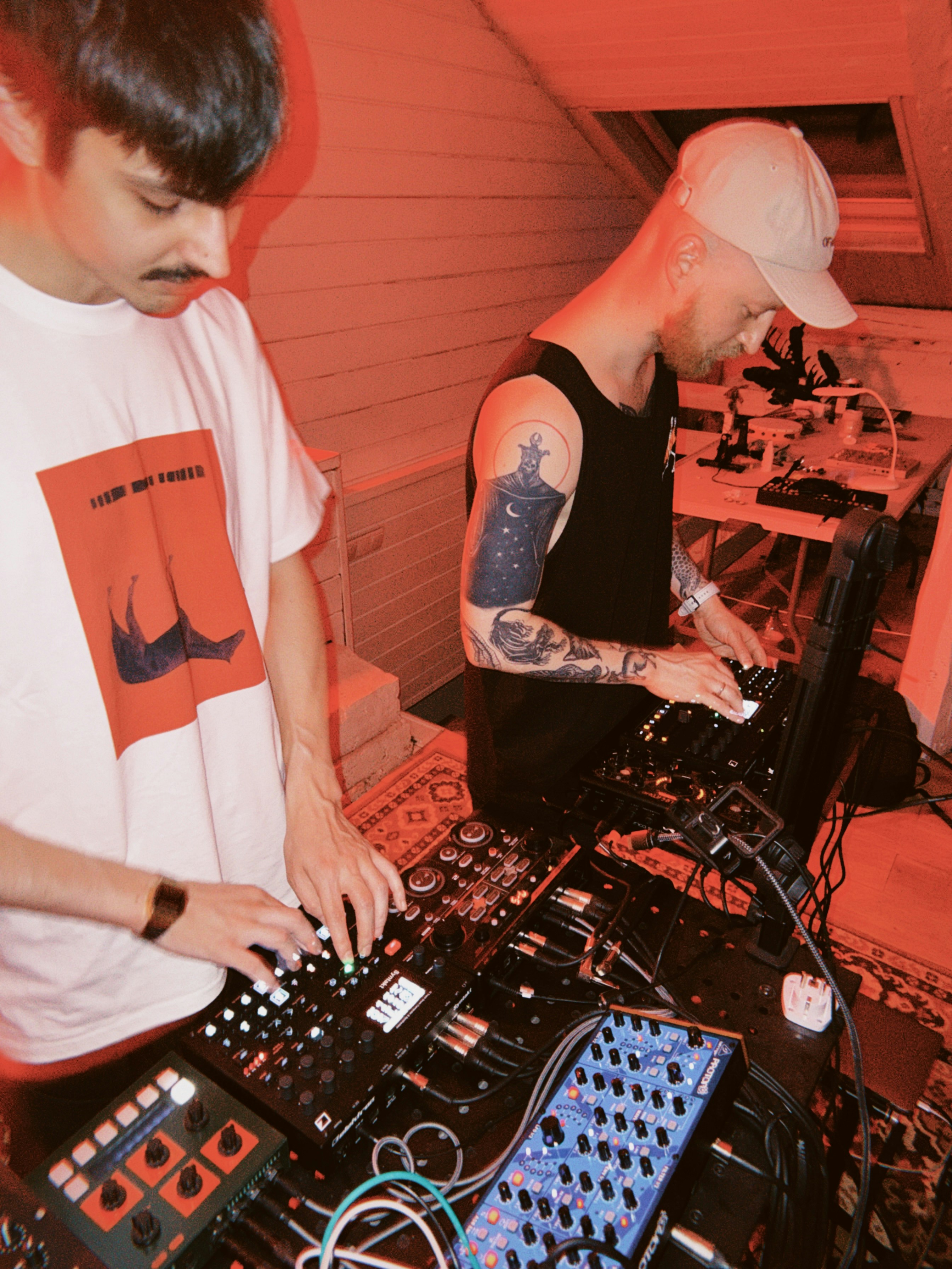 Two musicians operate a crowded DJ setup in a red-lit room, focused on mixing gear and controllers.