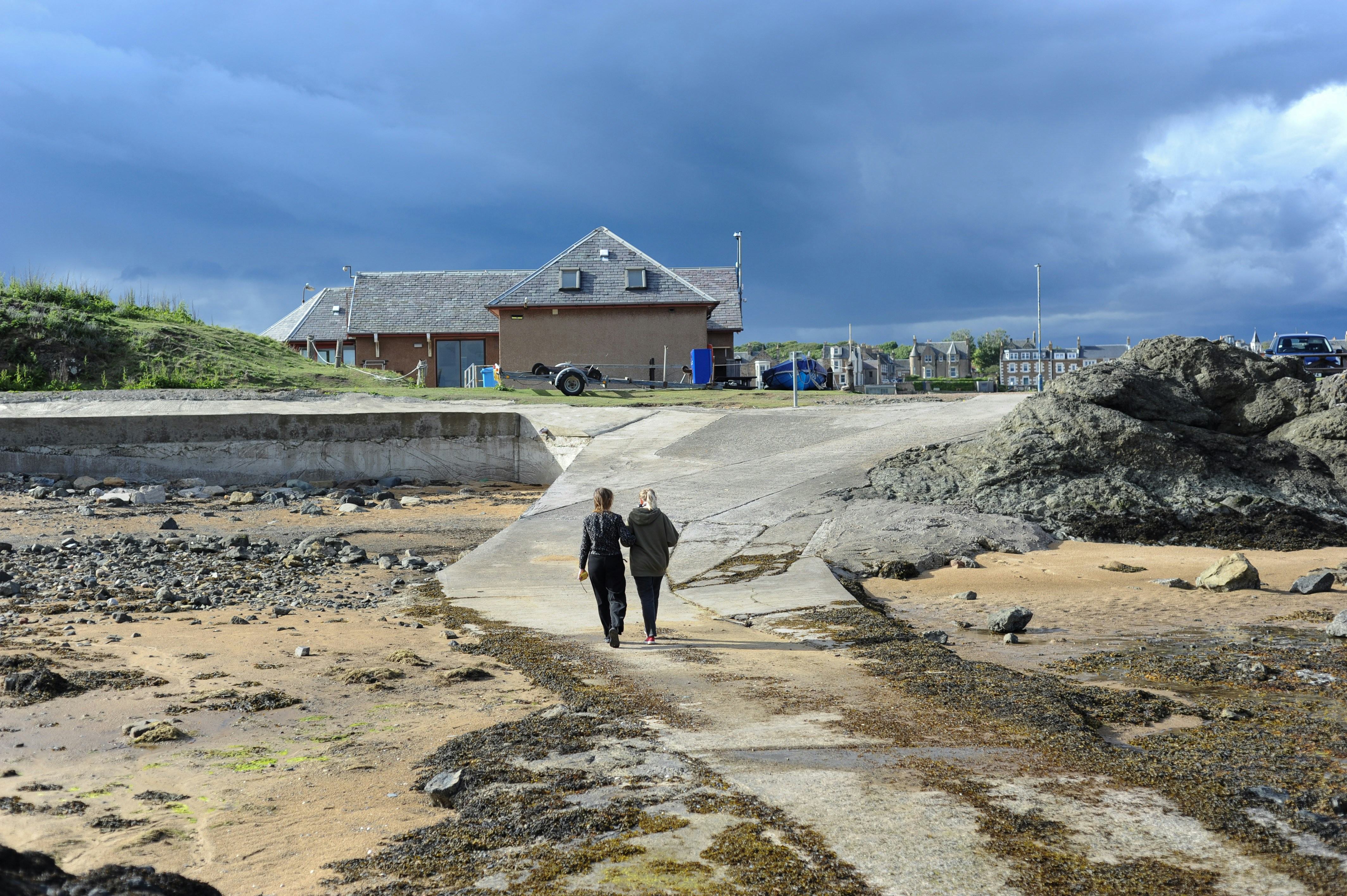 Two Travelers Crossing a Rocky Breakwater Toward a Quiet Village