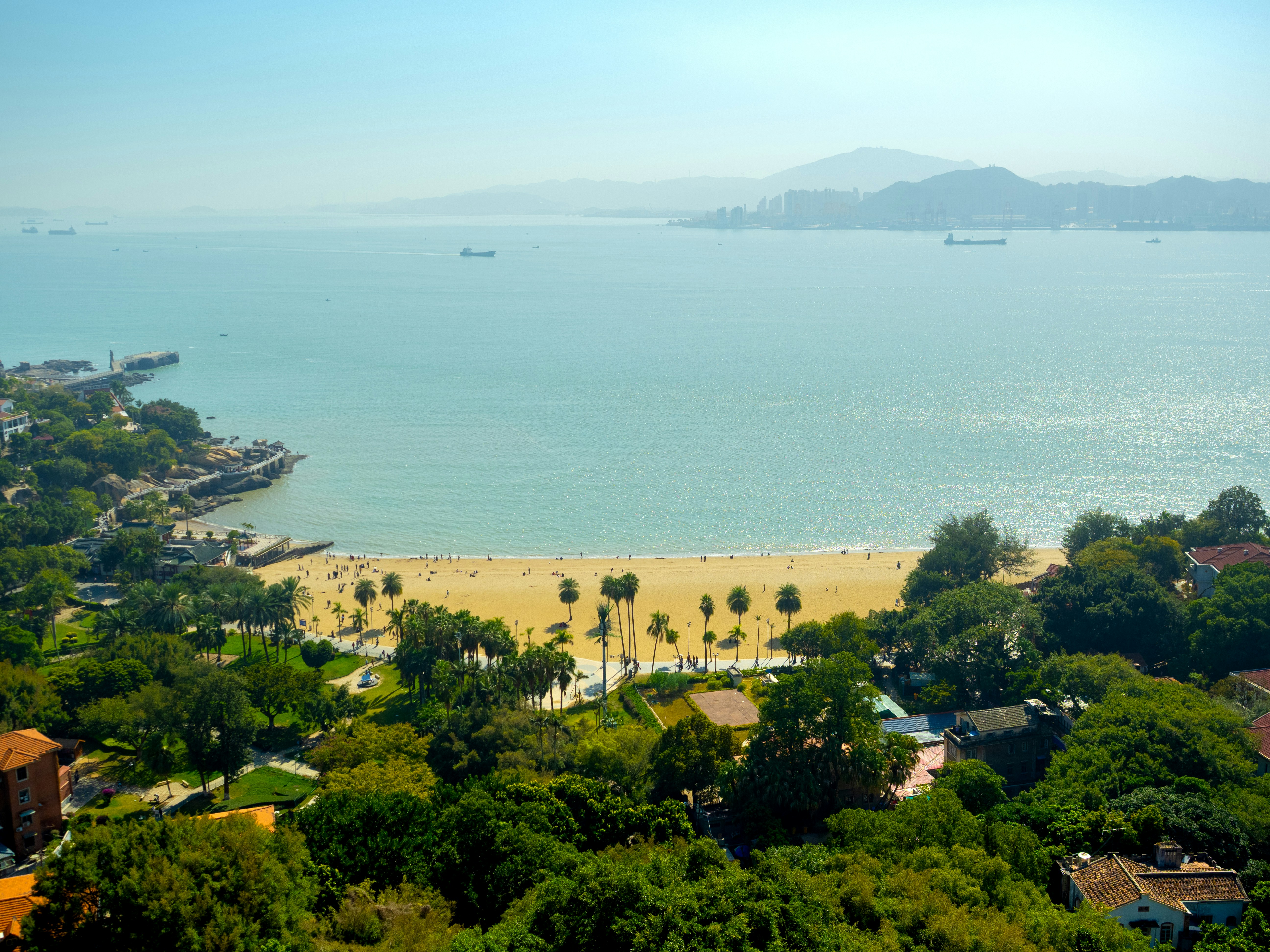 Coastal view with a sandy beach, turquoise water, and palm-lined foreground residential area.