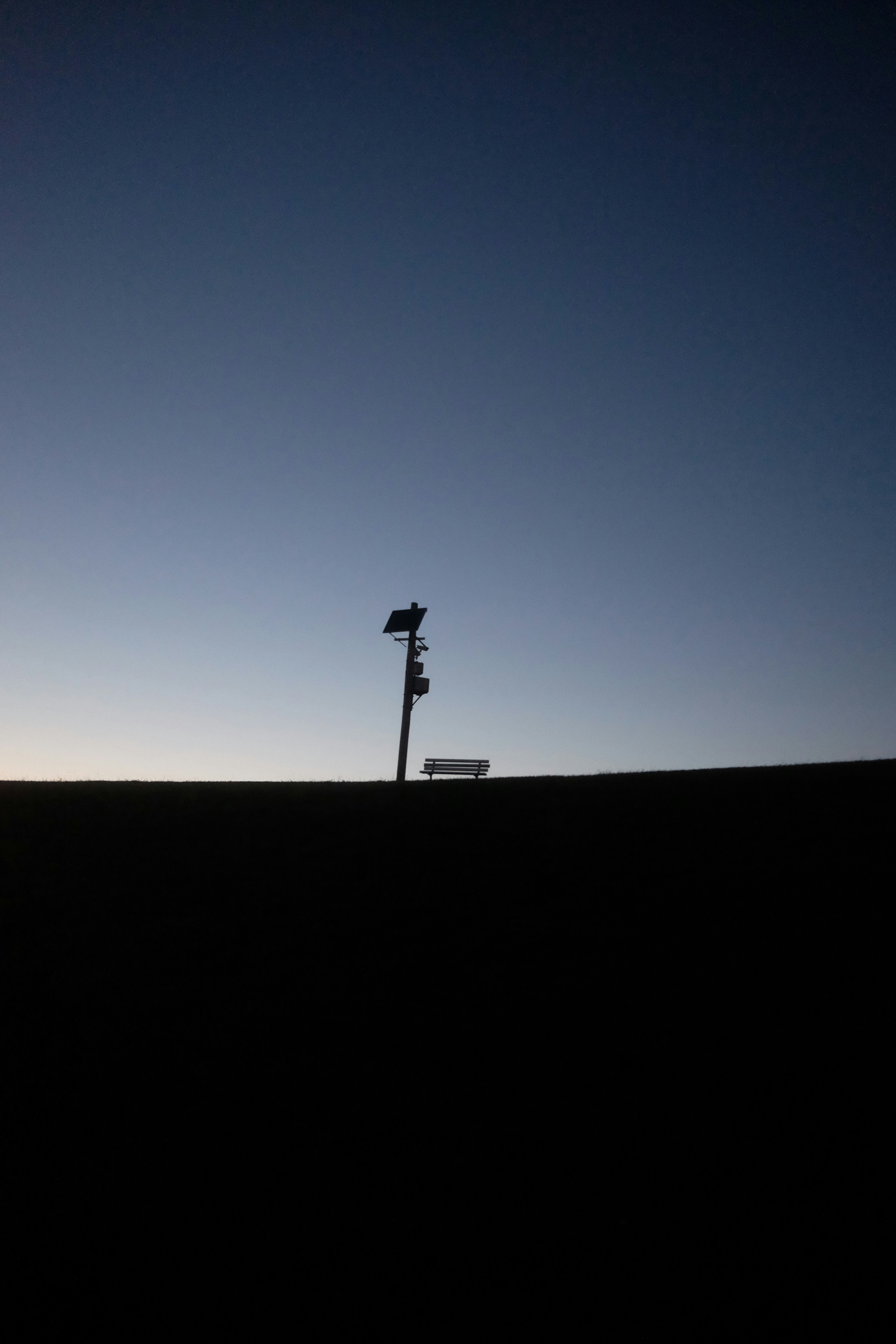 Vast gradient sky silhouettes a lone bench beside a signpost on a gentle horizon line, creating a quiet, contemplative scene.