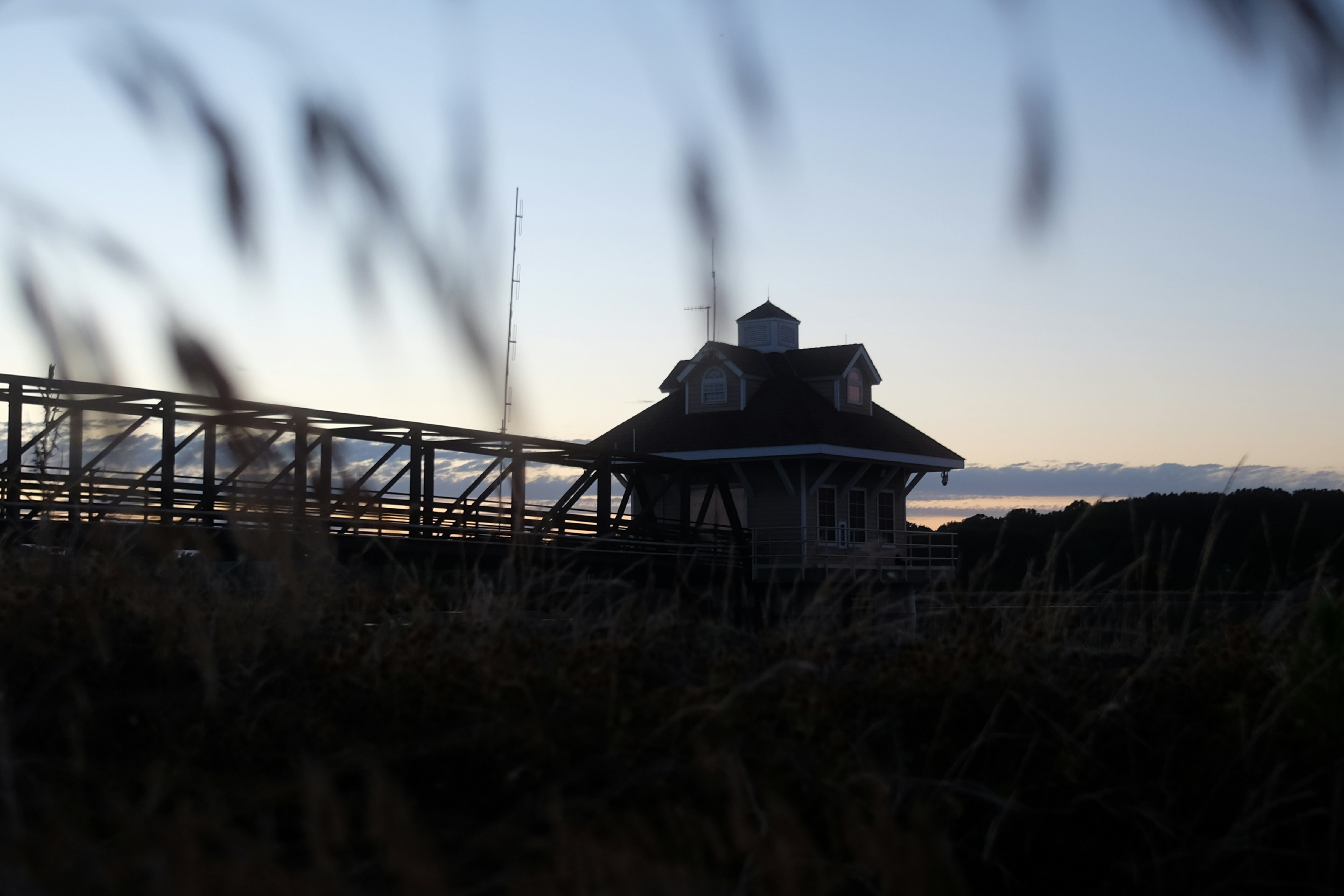 Dusk light silhouettes a small pier house at the end of a wooden walkway, with blurred grasses in the foreground and a calm horizon in the background. A logo-like structure stands as the focal point of the scene.