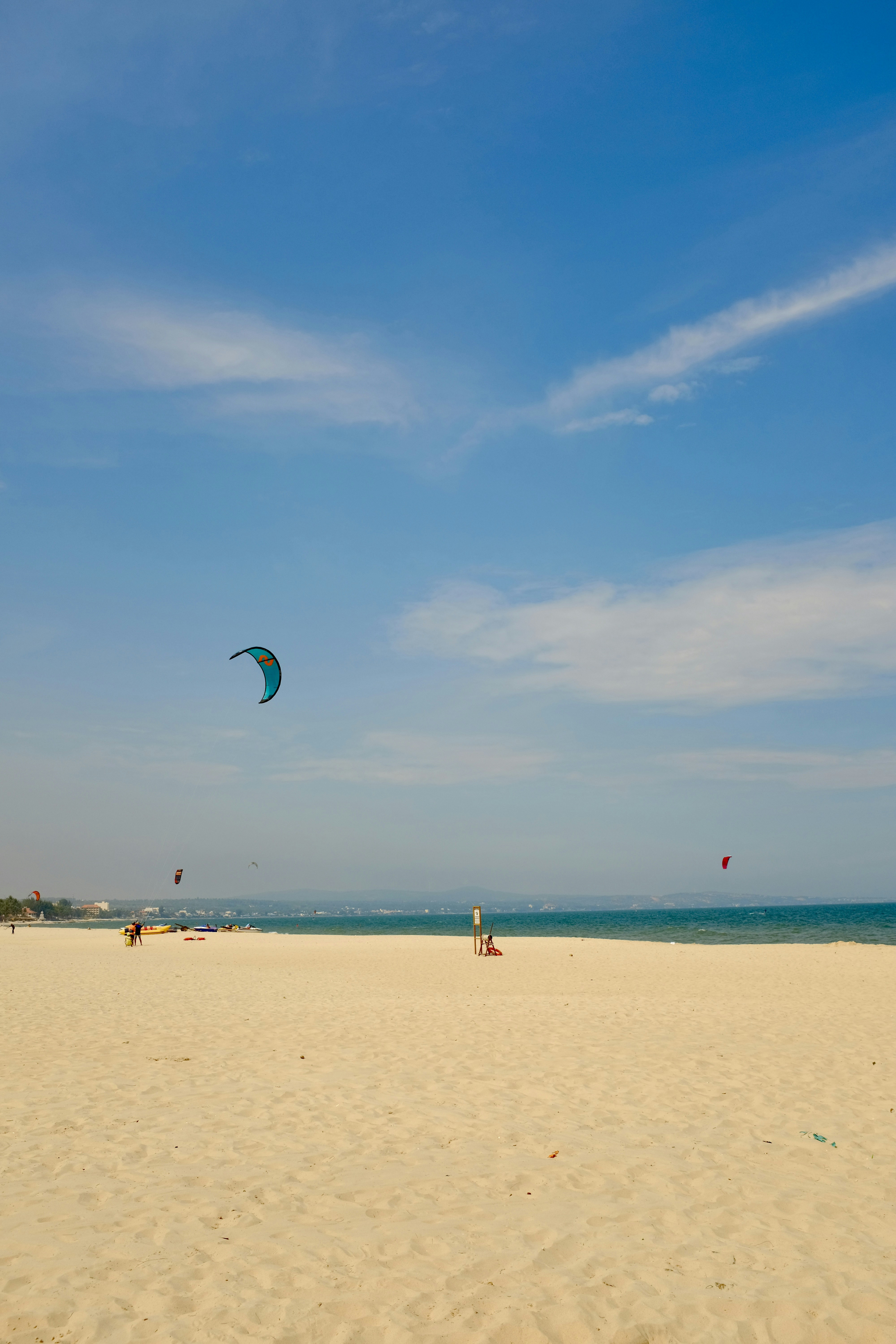 Sunlit beach with pale sand meeting the calm blue sea under a clear sky; small kite surfers pinpoints along the horizon, emphasizing spaciousness of the coastline.