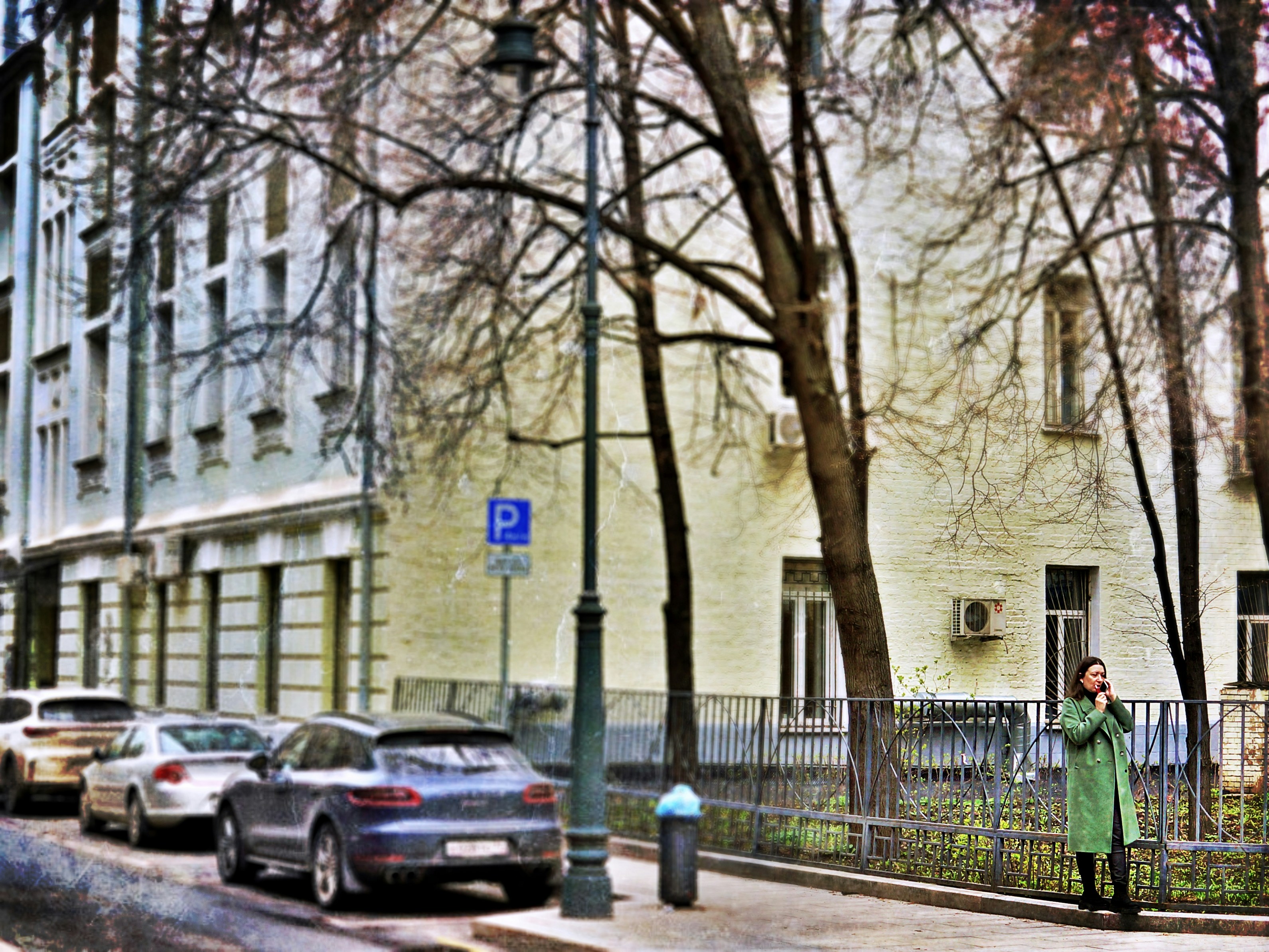 Woman on a city sidewalk framed by bare trees and parked cars