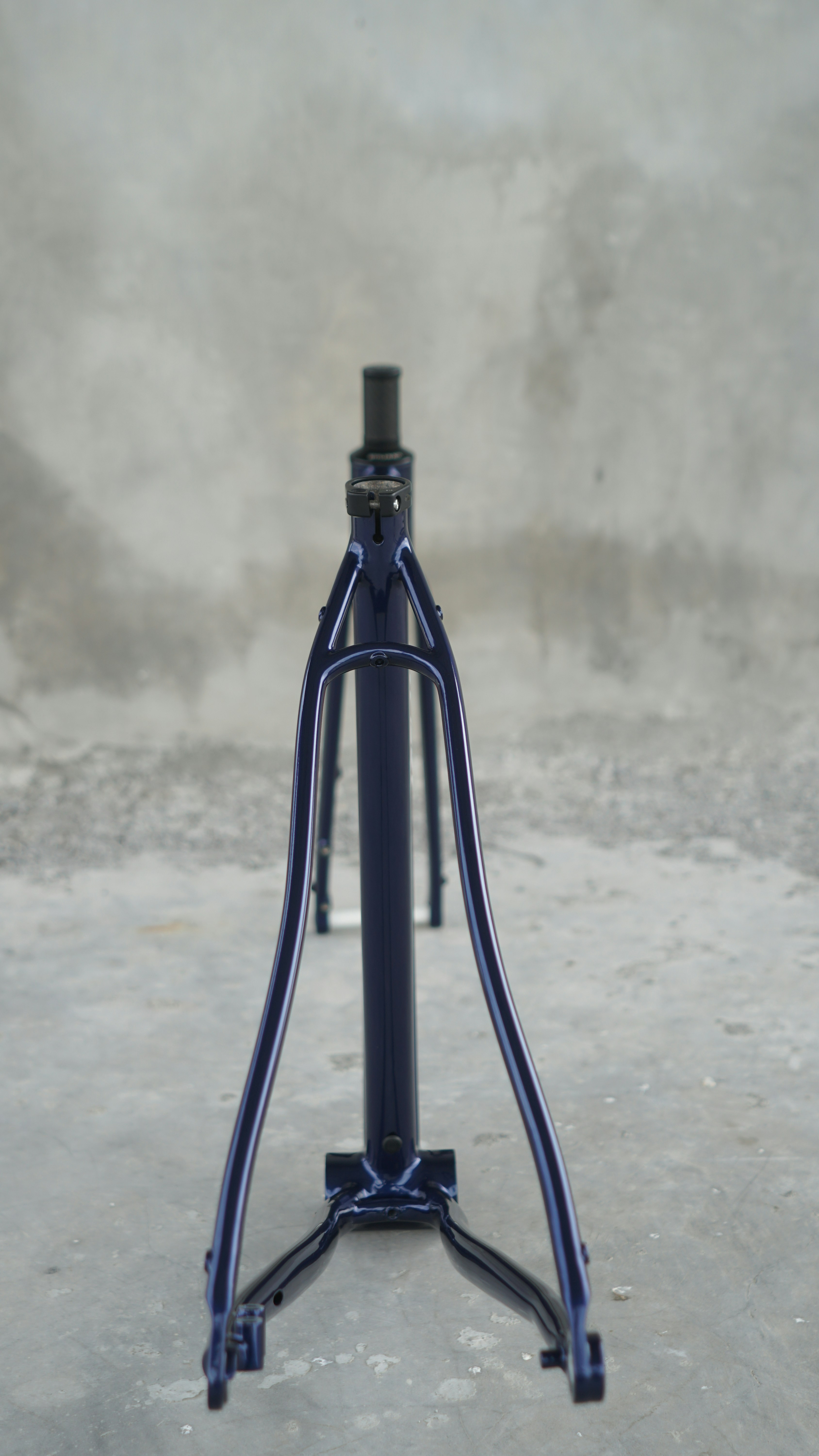 Blue bicycle fork stands centered against a muted concrete backdrop with shallow depth of field, emphasizing the metallic frame. Photograph of a bicycle fork structure, non-illustrative.