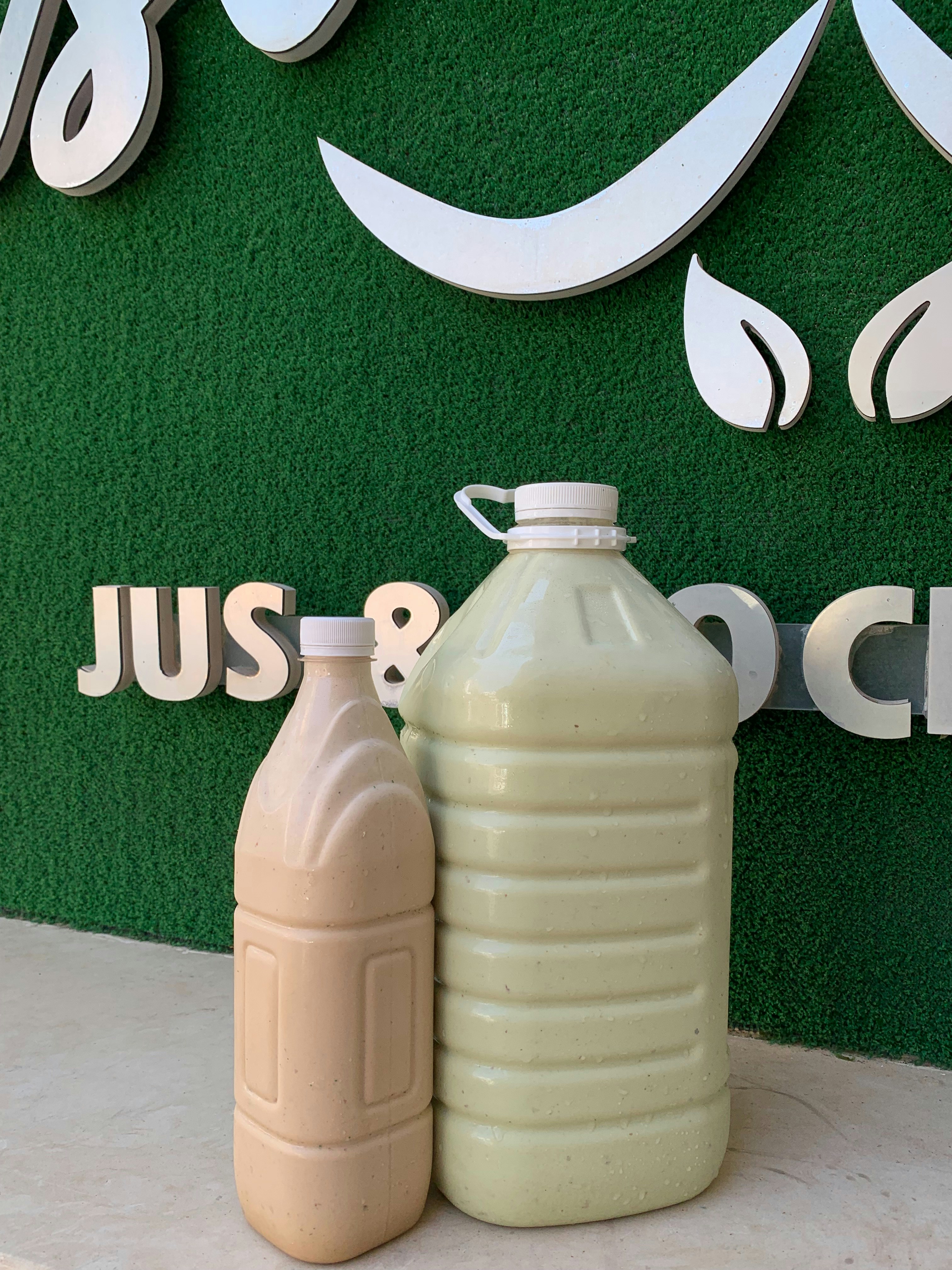 Two plastic jugs filled with liquids sit on a concrete surface in front of a green, textured wall with partial white logo lettering.