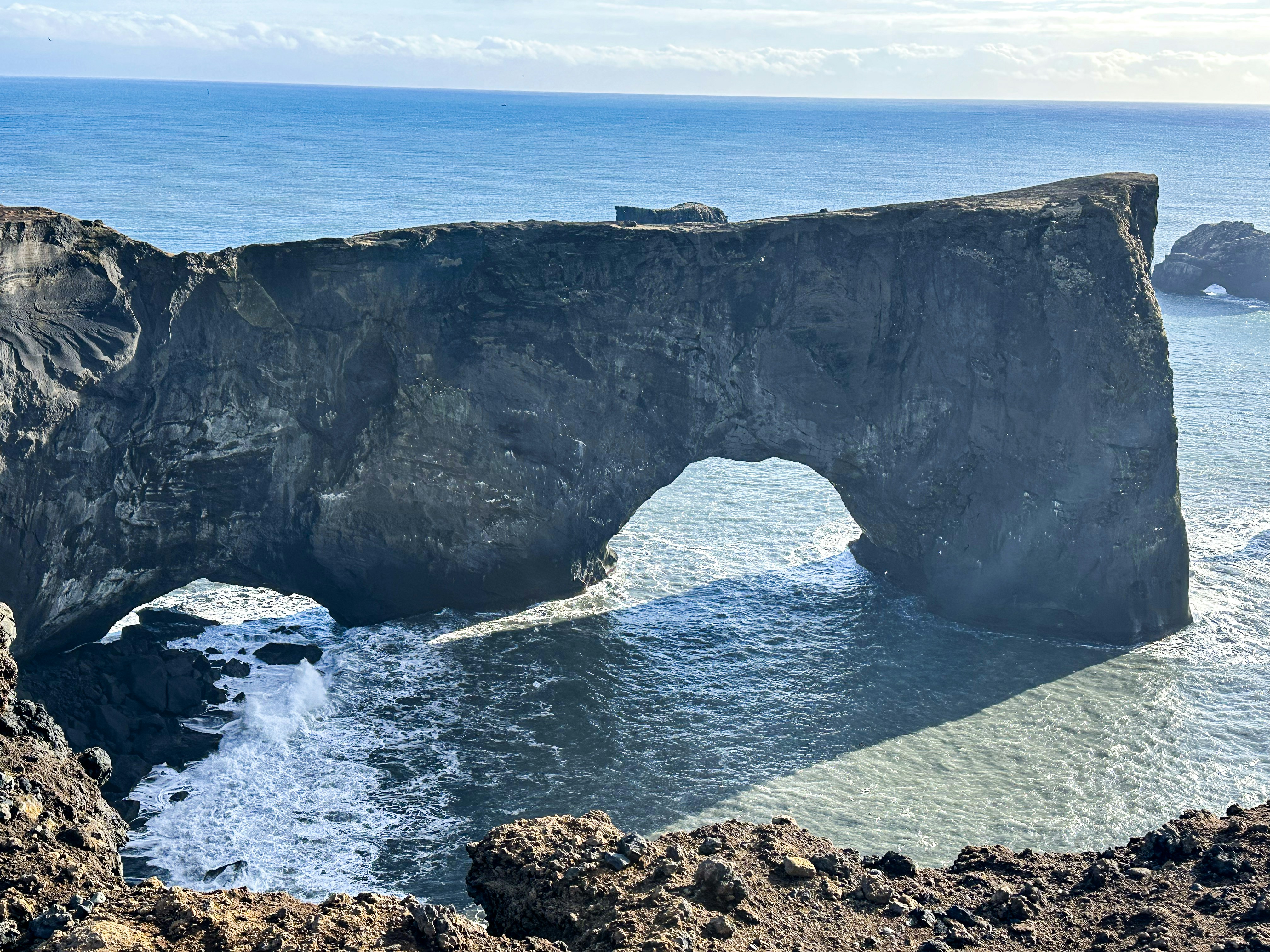 Sea arch carved in dark basalt stands against blue water, with rocky foreground and sunlight casting a shadow inside the arch.