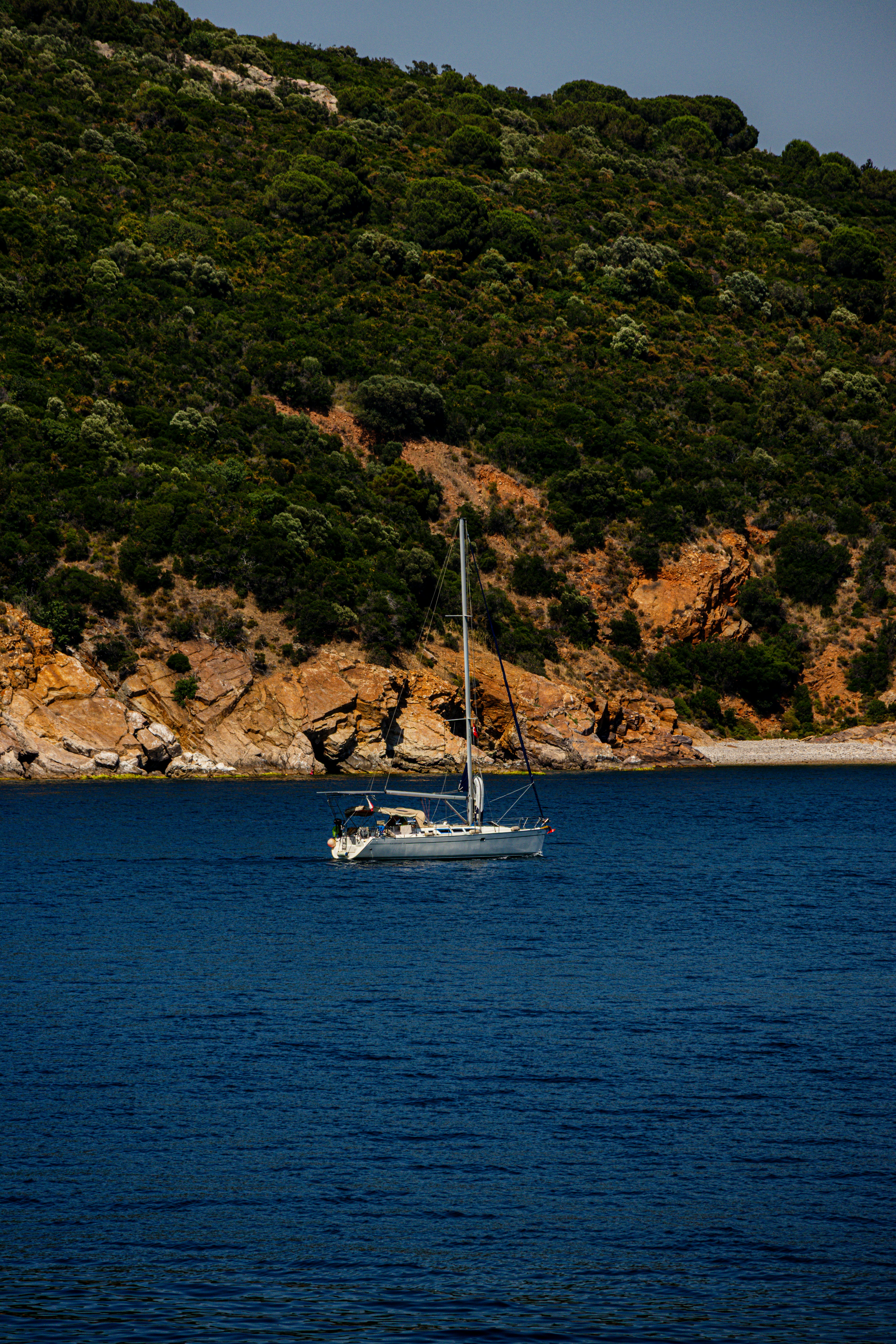 Sailboat rests on deep blue water with rugged, sunlit cliffs and green shrubbery in the background.