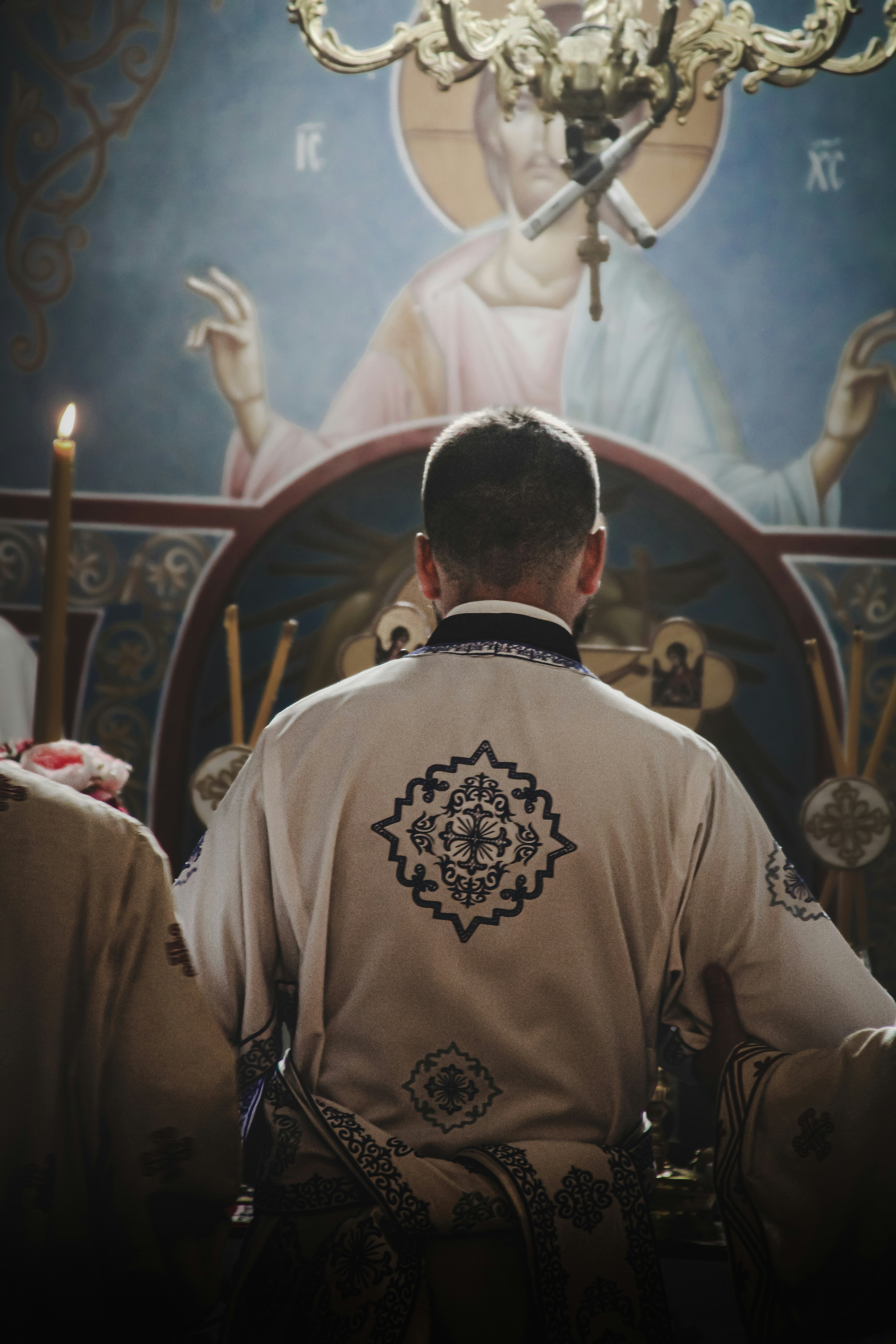 Cleric facing an illuminated icon during a ritual, backlit by candlelight; ornate embroidered robes and sacred symbols frame the scene.