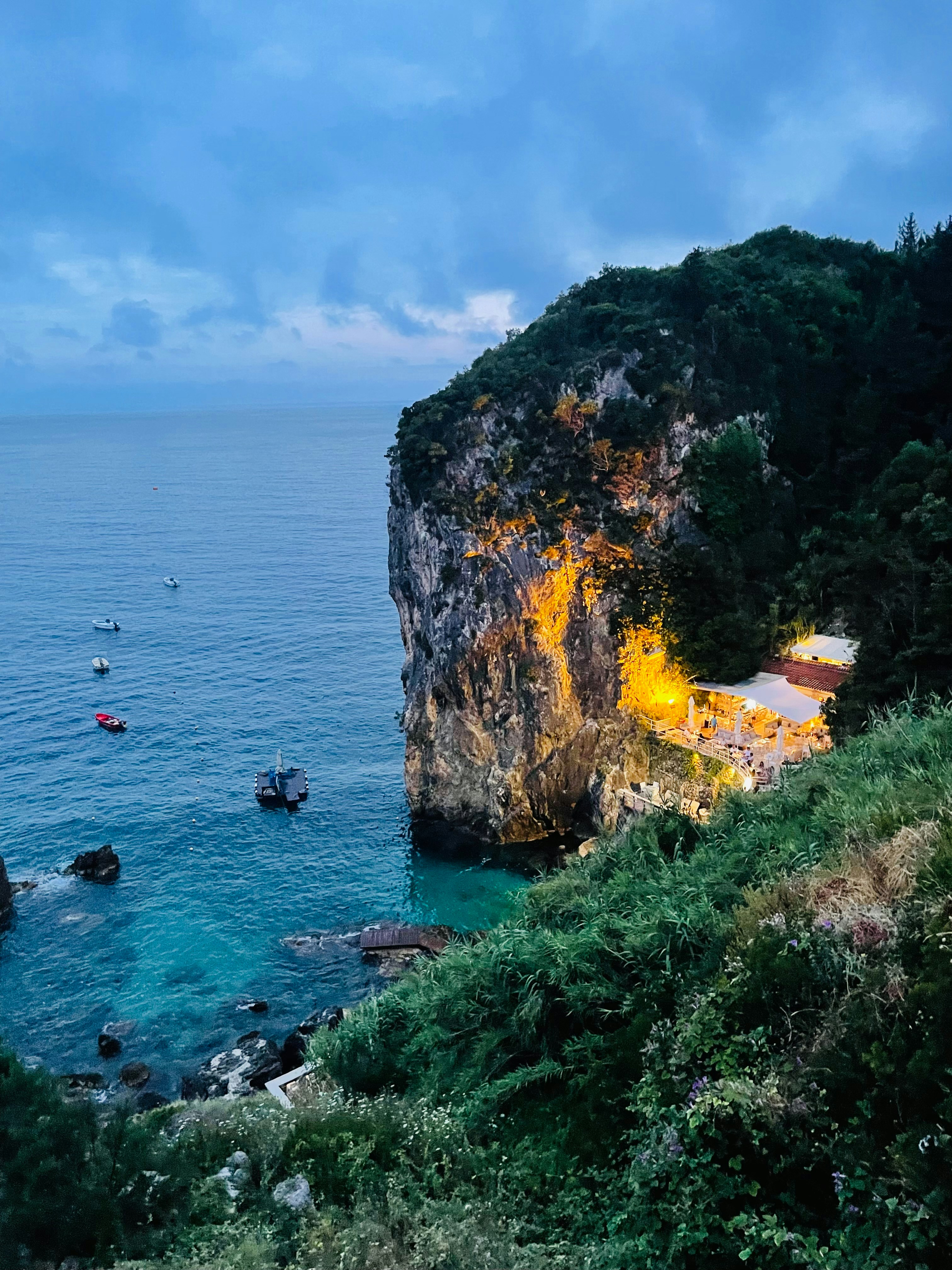 Steep rocky cliff meets turquoise sea with a lit terrace perched along the coastline, framed by lush greenery and a calm horizon.
