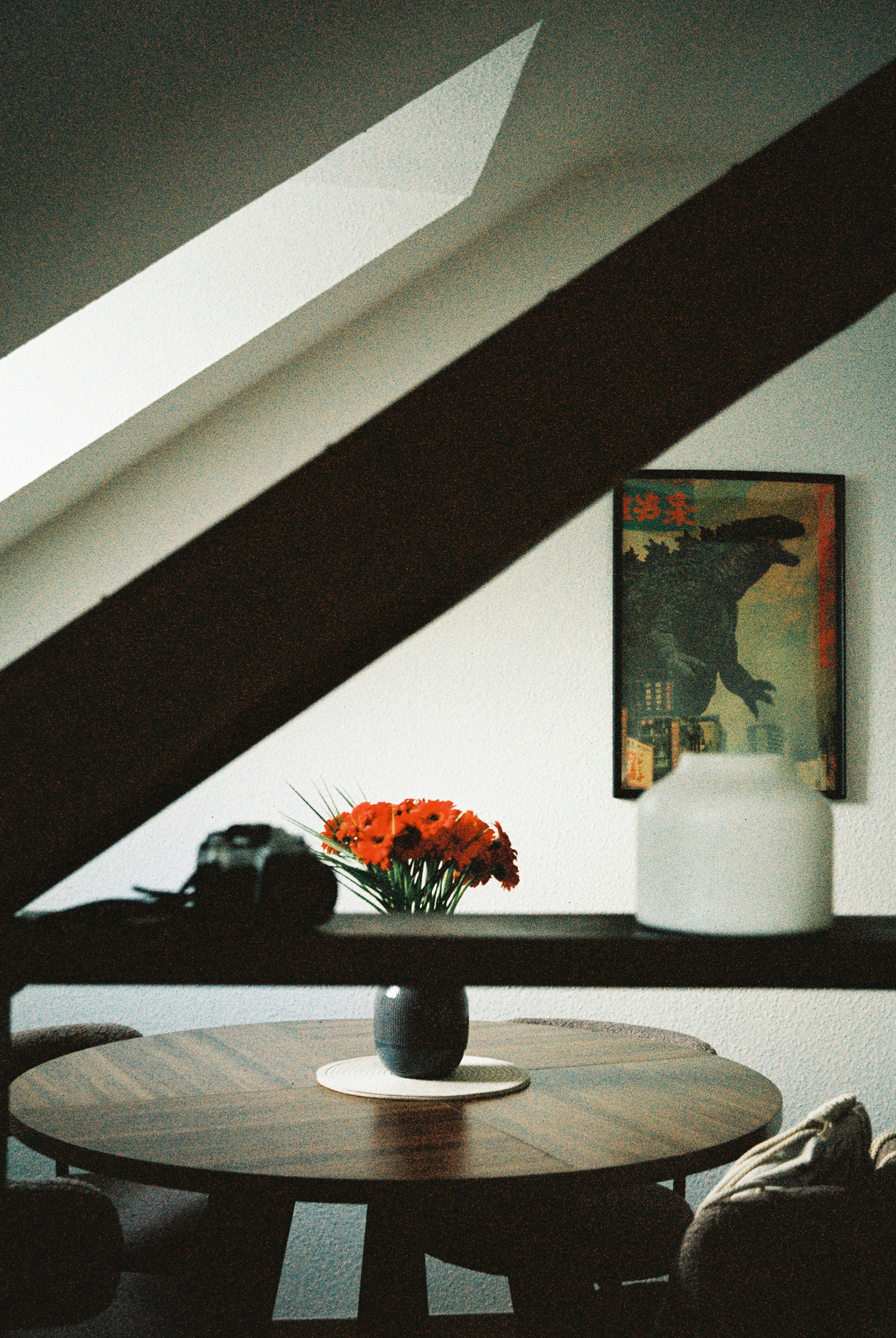Interior still life with a circular wooden table, vase of orange flowers, a camera and a framed artwork on a wall under diagonal architectural beams.
