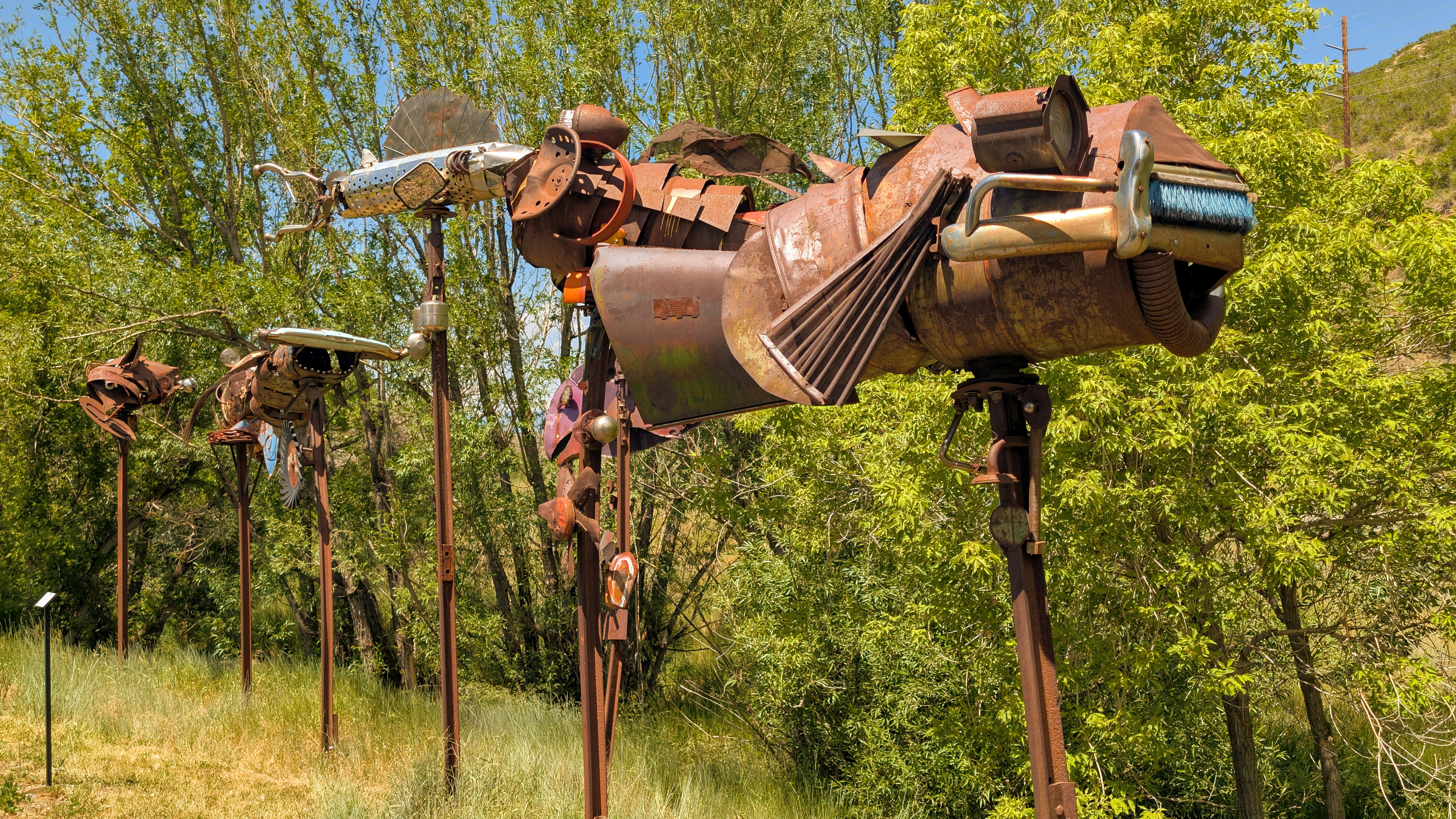 Rust-colored metal sculptures on tripods rise through a sunlit grove, forming an outdoor art installation amidst green trees and dry grass. A weathered machinery aesthetic dominates the scene.