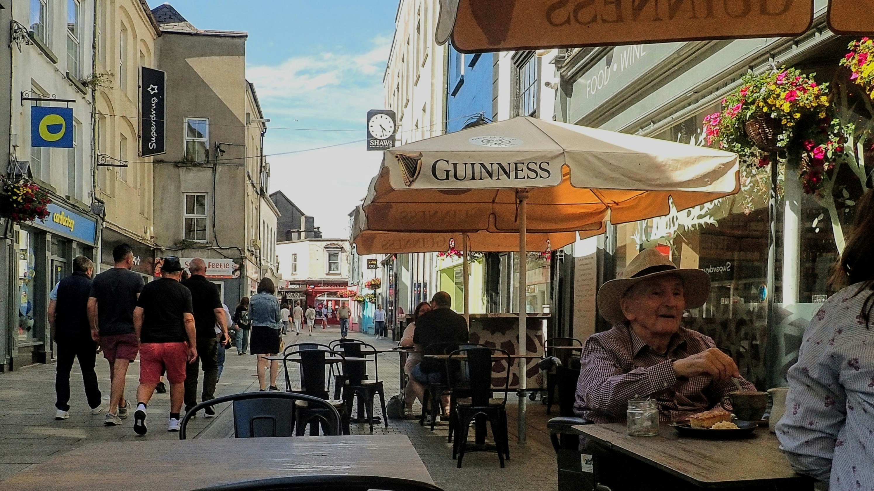 Sunlit street café scene with patrons at outdoor tables and a shopfront on a pedestrian shopping lane.