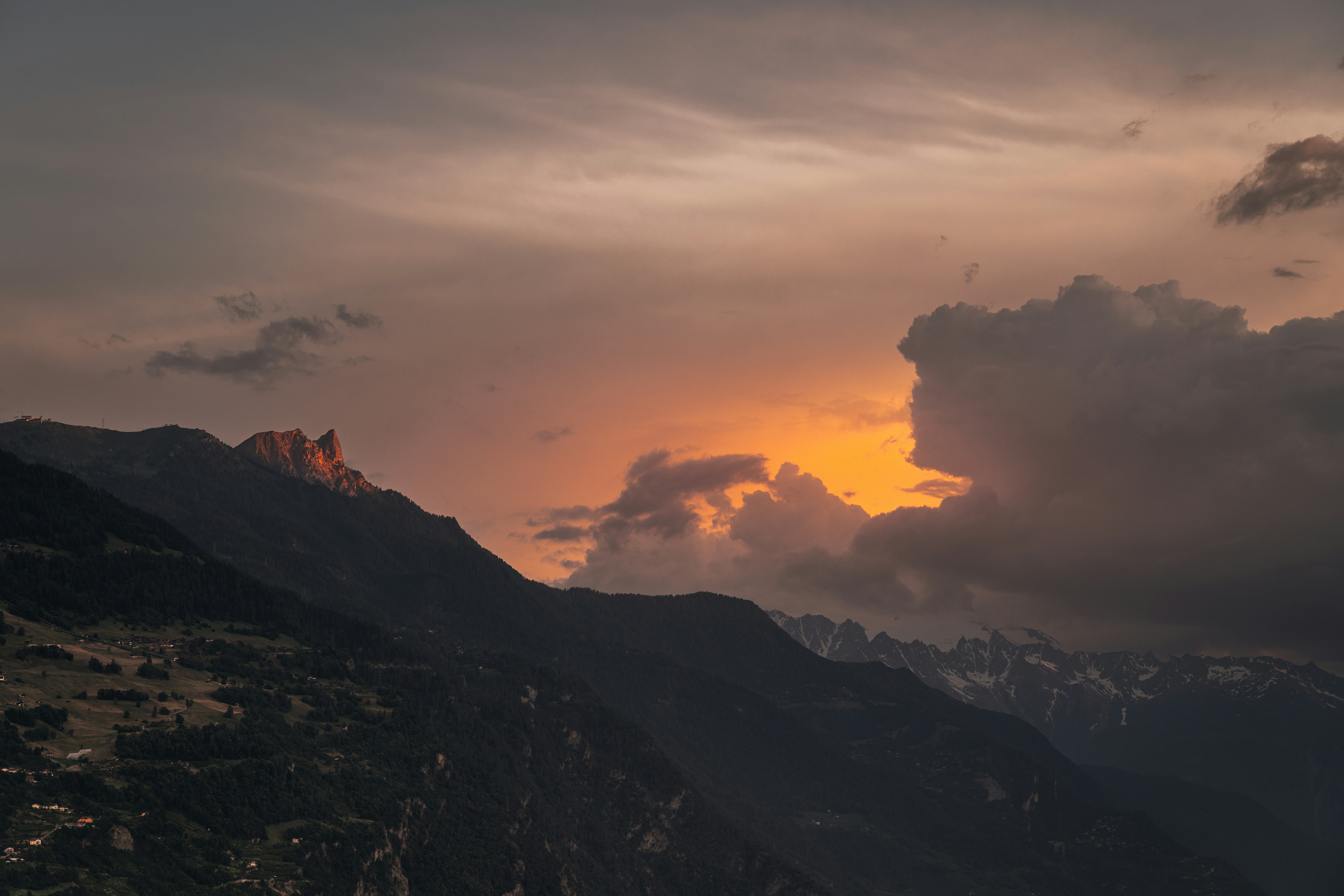 Sunset ignites the jagged summit silhouettes over rolling valleys, with warm light brushing the ridge as clouds gather above. A landscape study emphasizing mood and depth.