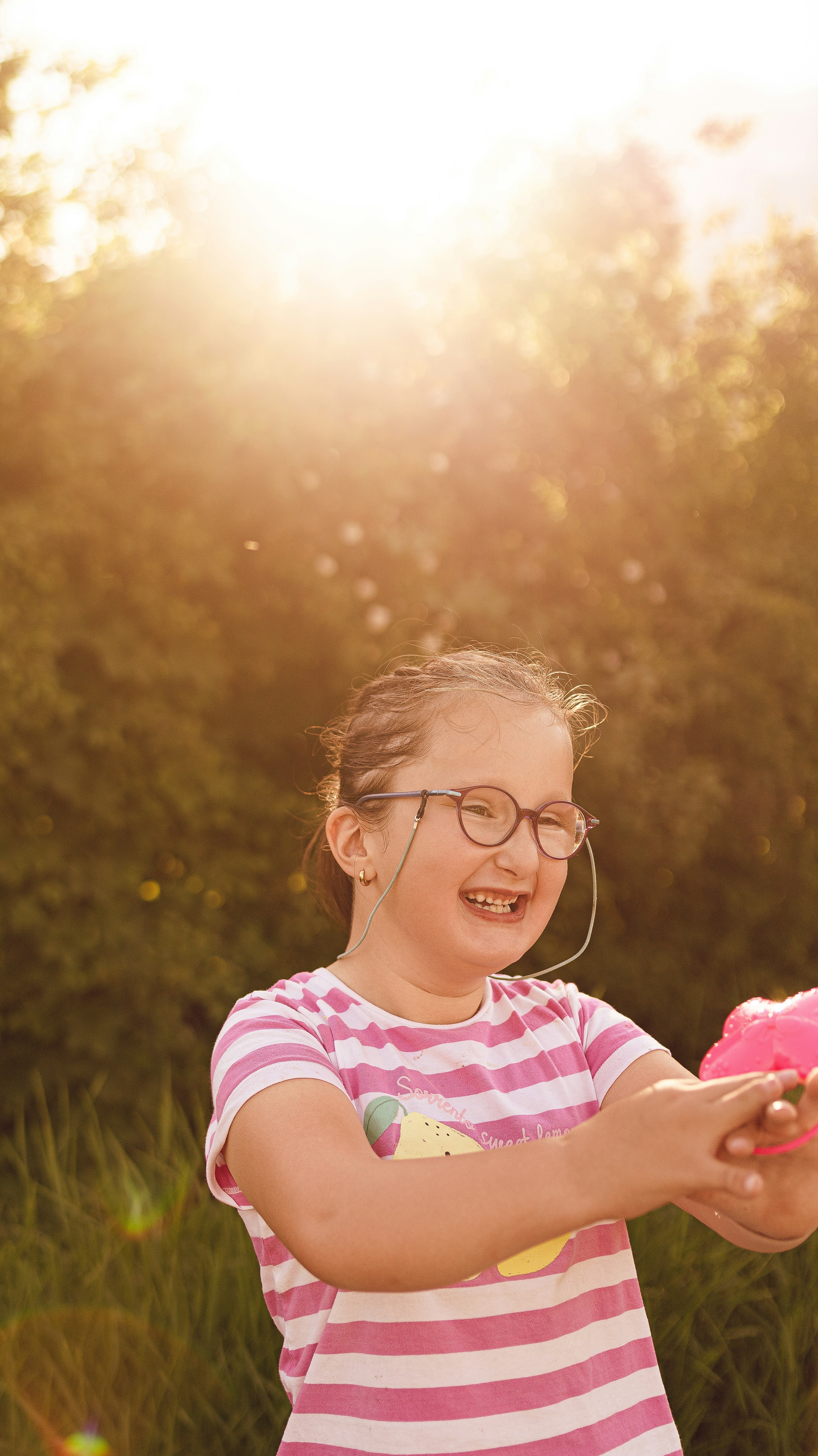 Young girl with glasses laughs as she extends her arms toward a pink toy, bathed in warm sunset light.