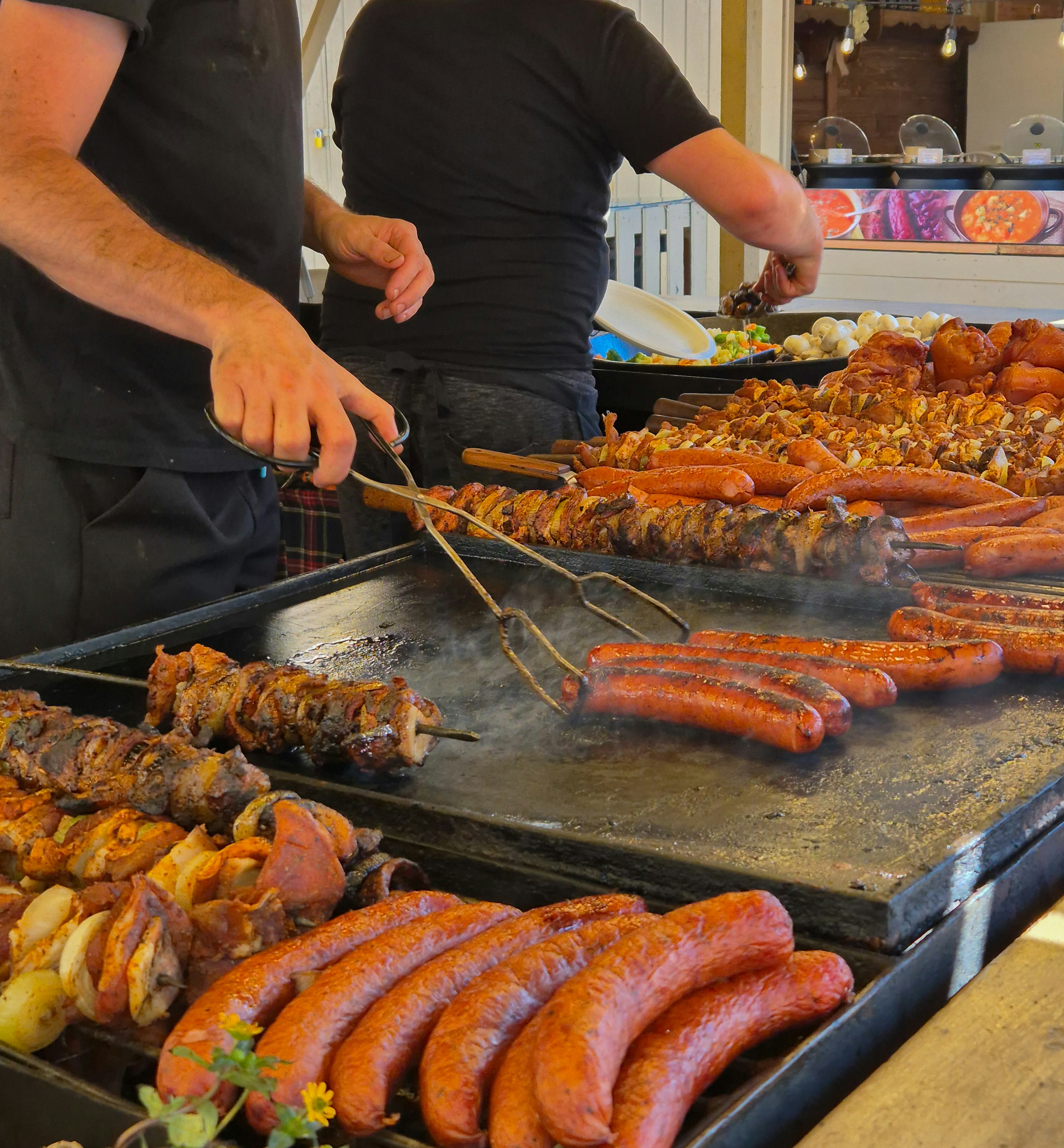 Vibrant street-market grill scene with an array of sausages and skewers cooking on a wide flat grate amid busy vendors.