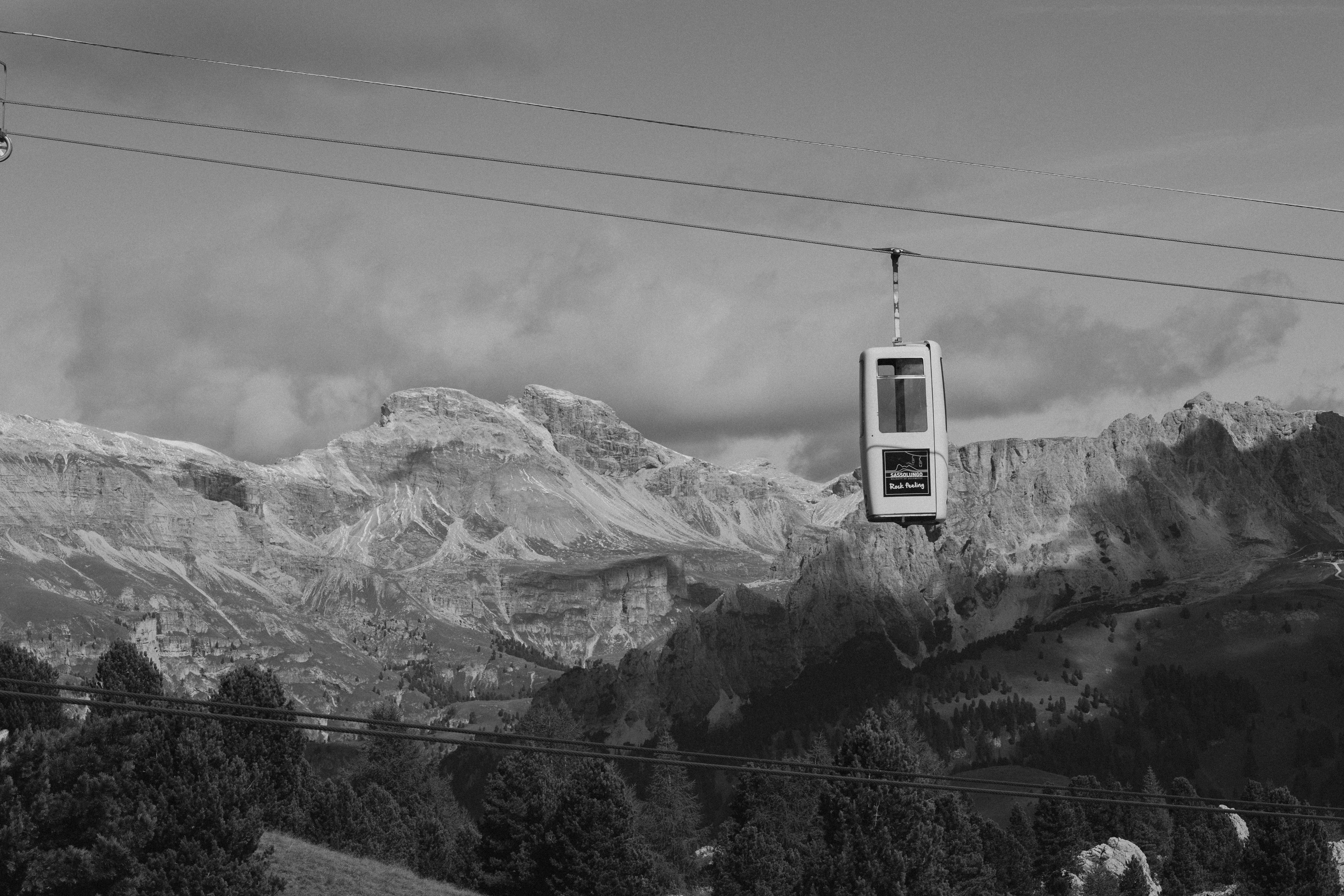 Monochrome scene of a cable car gliding above a forested valley with jagged mountain peaks in the background.