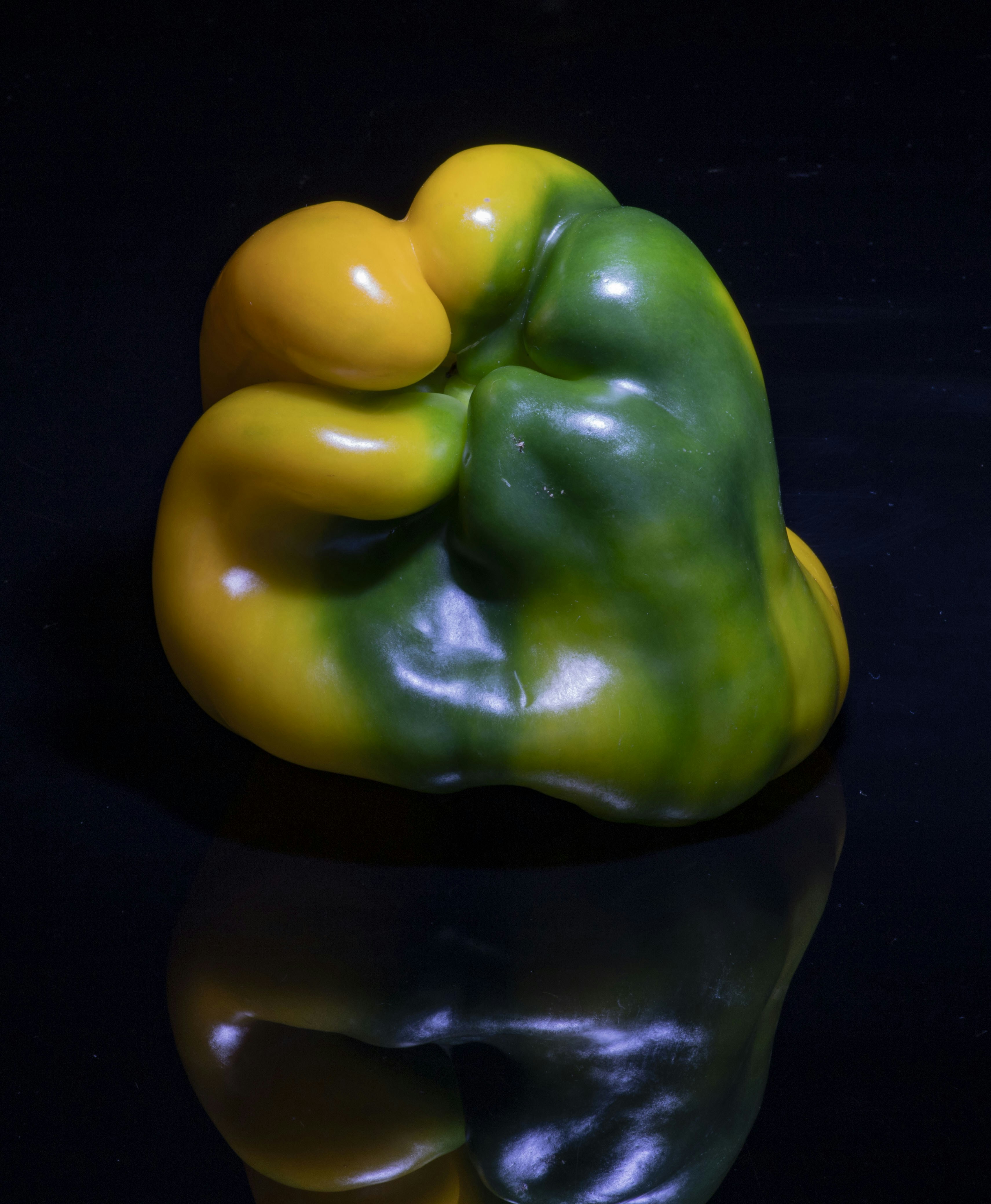 Glossy bell pepper resting on a dark reflective surface, illuminated with cool highlights and warm yellows-green tones. A studio still-life emphasizing color, curvature, and texture without background distraction.