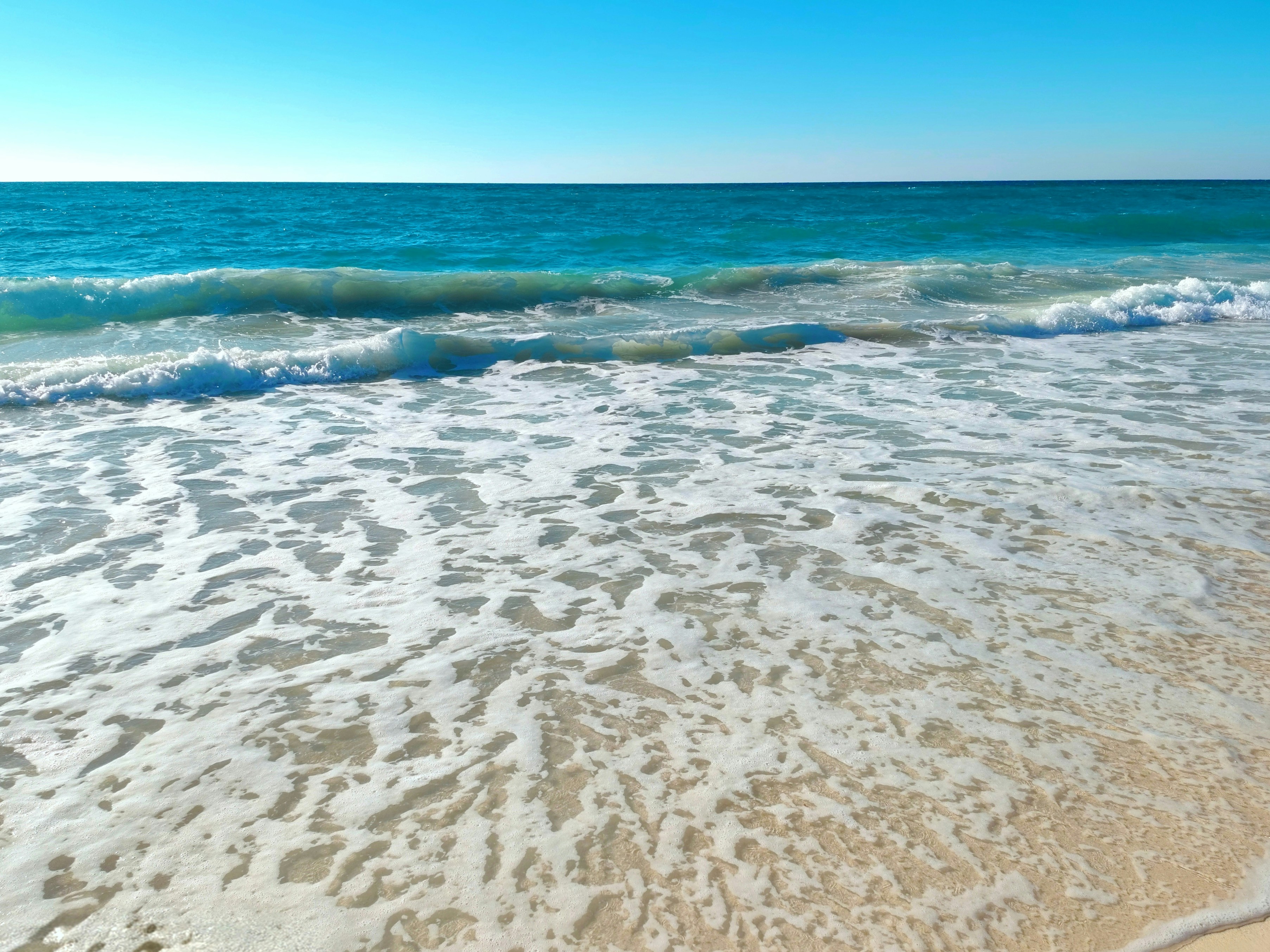 Sunlit shoreline with frothy waves meeting a sandy beach under a clear blue sky; a serene seascape highlighting shallow, patterned foam and turquoise water. (photograph) Real-world beach scene.