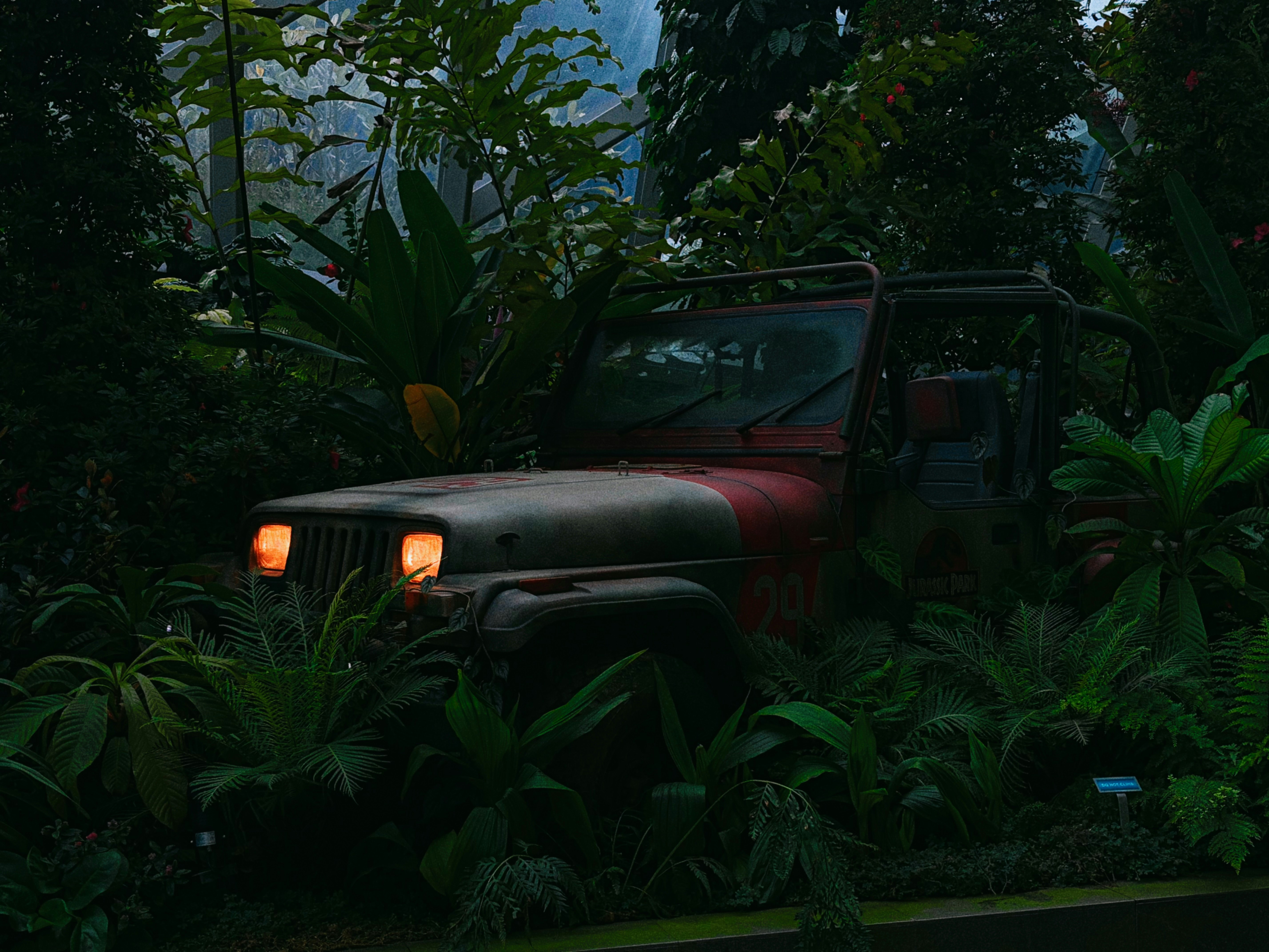 Red jeep partially hidden among dense tropical foliage in a dim garden scene with muted headlights glowing.