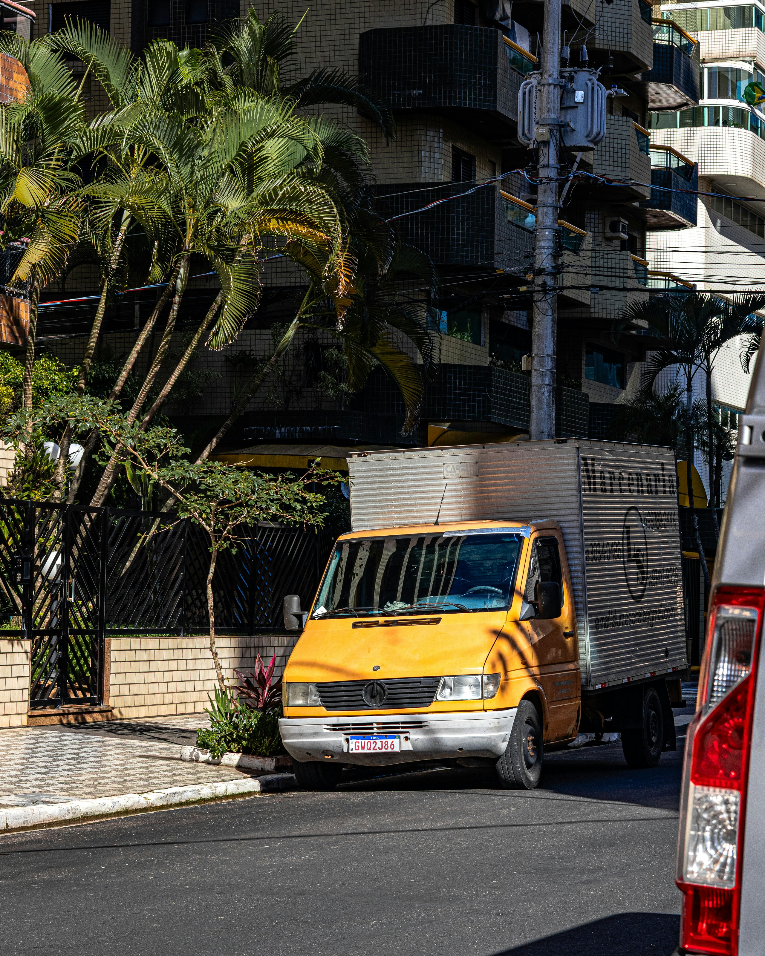 A yellow delivery truck is parked on a sunny street in front of a modern building with palm trees.