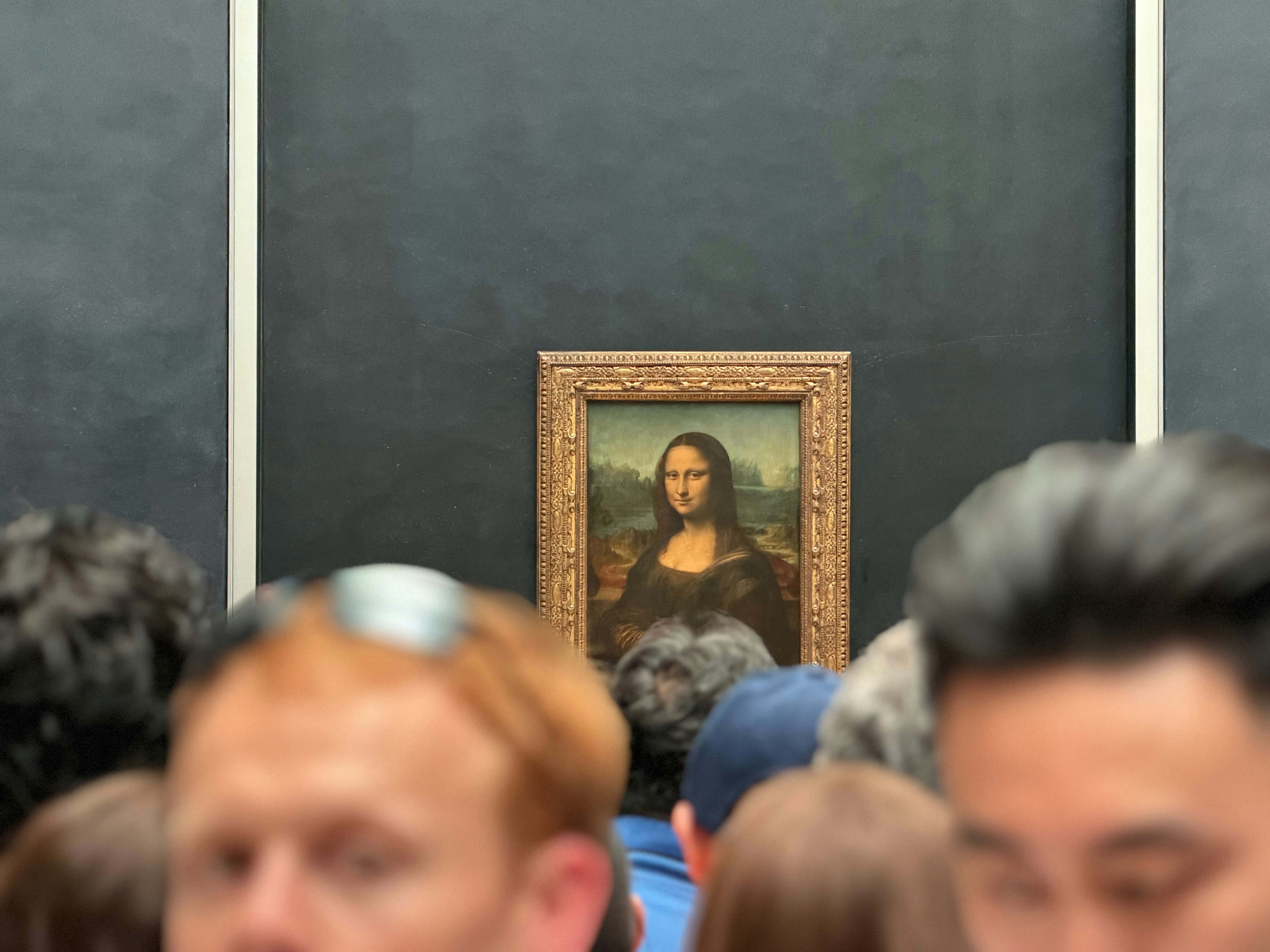 A crowded gallery scene with a small, framed Mona Lisa drawing centered on a dark wall, drawing attention amid blurred visitors.
