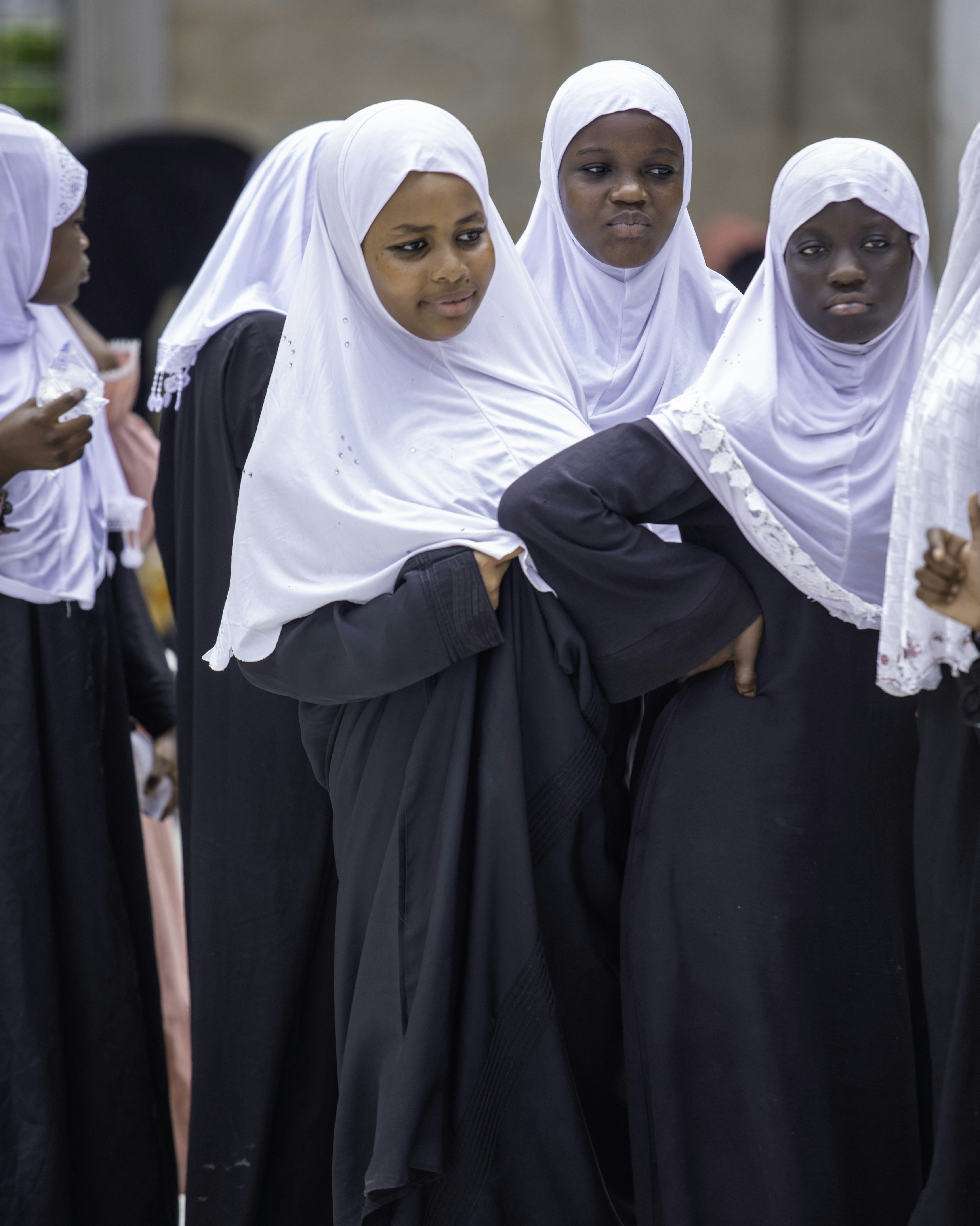 Group of young women in white hijabs and black dresses stand together at a cultural event, expressions attentive and calm. A portrait-focused scene with emphasis on head coverings and modest attire.