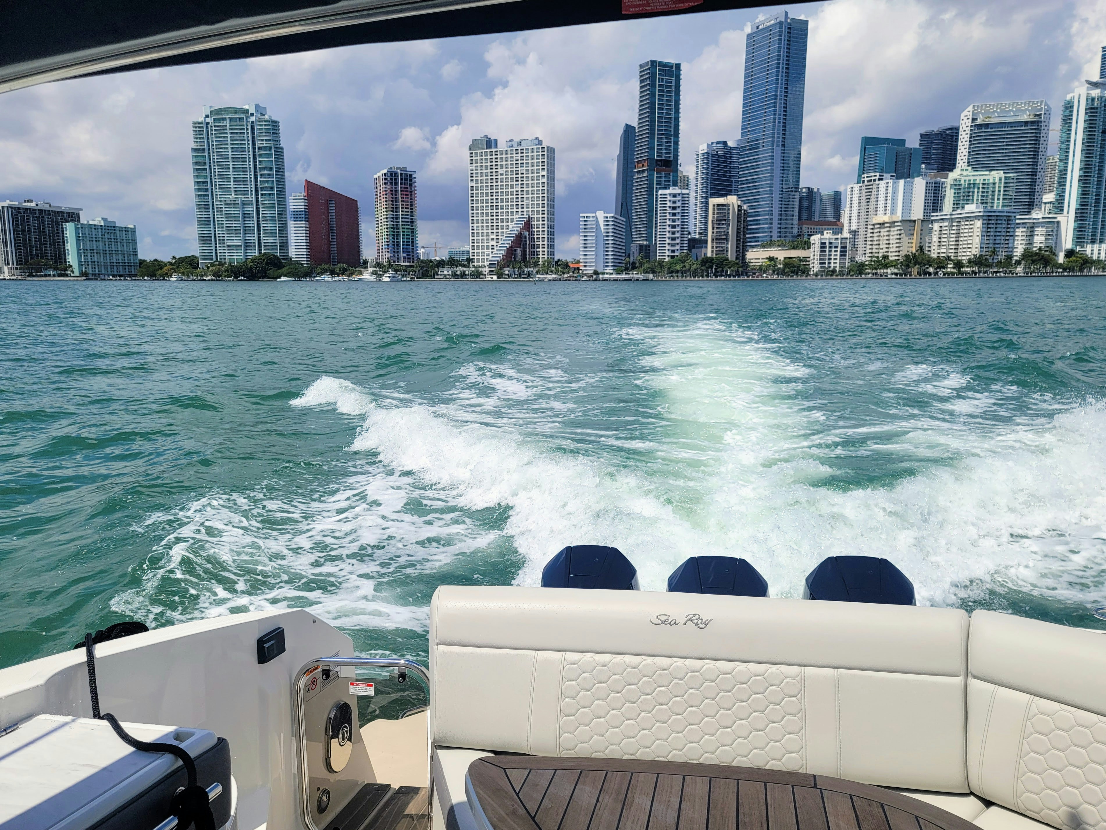 Waterfront skyline viewed from a speeding boat with wake in the foreground; boats’ seating and ocean spray frame the urban horizon.