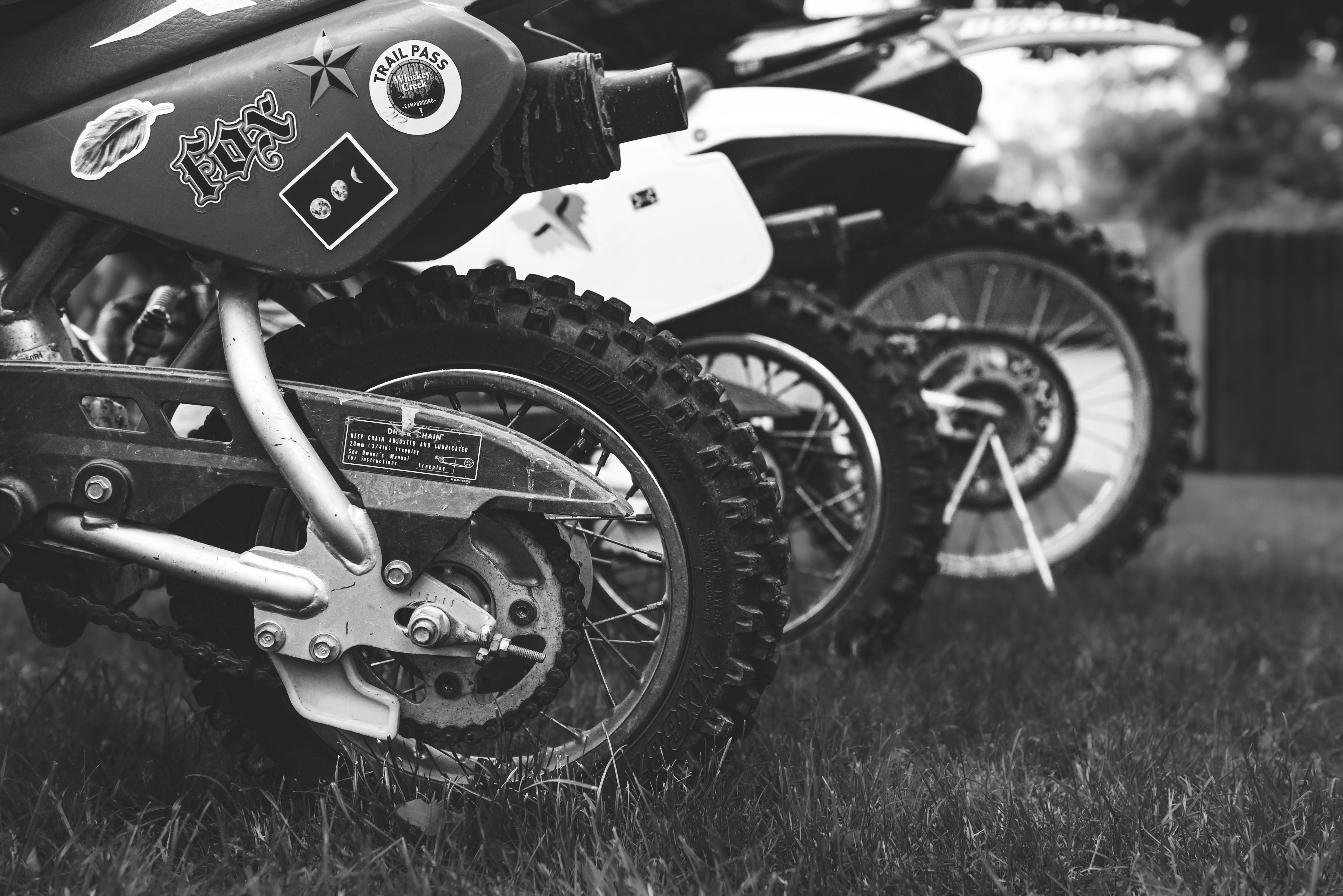 A row of dirt bikes parked on grass, showing knobby tires and chain details with stickers on the bodywork.
