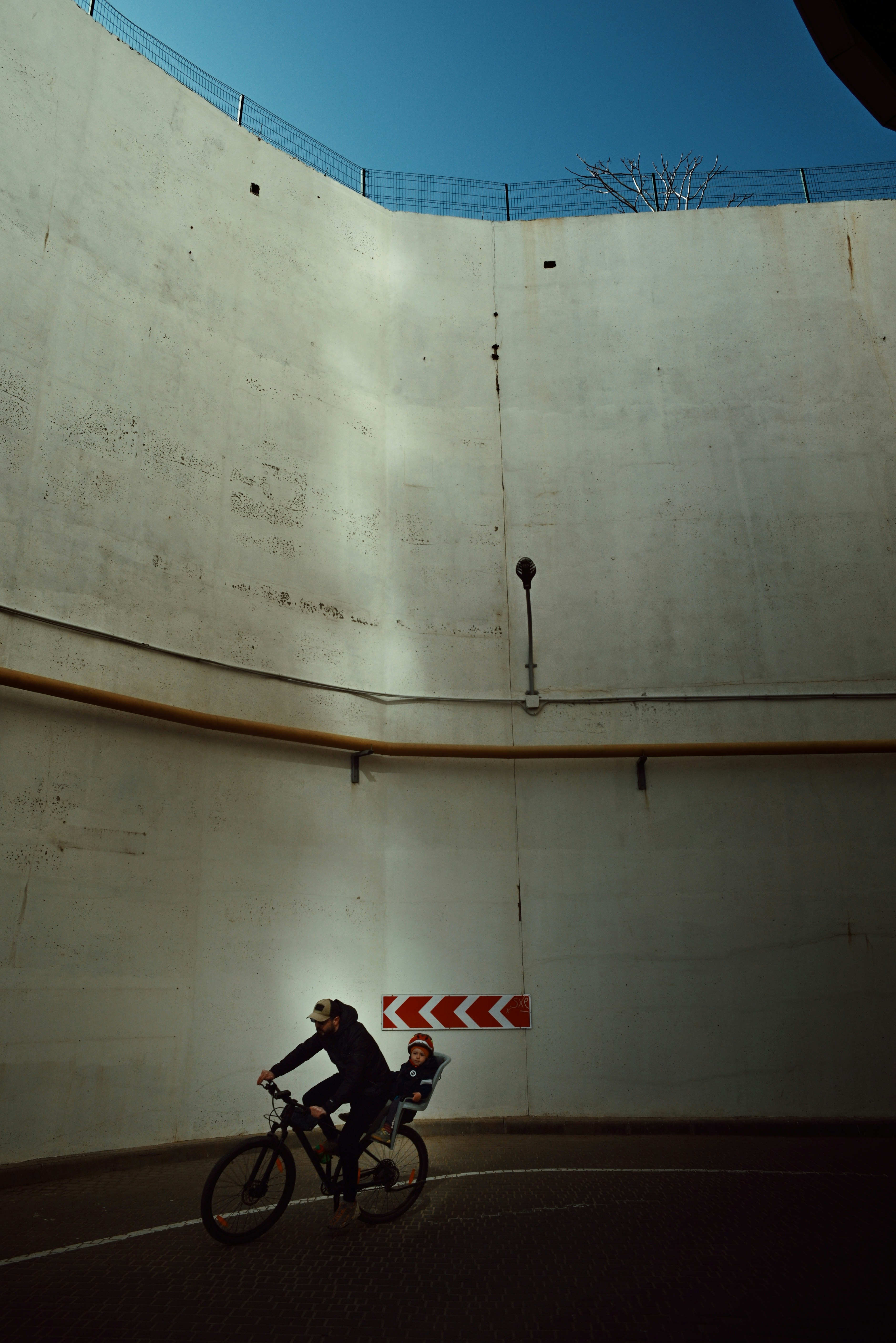 A dramatic curve of a massive concrete retaining wall looms over a cyclist carrying a small child in a rear seat. Bathed in a striking pool of sunlight against the deep shadows of the underpass, the duo navigates the sharp bend, emphasized by a red-and-white chevron sign, while a clear blue sky peeks over the top edge.
