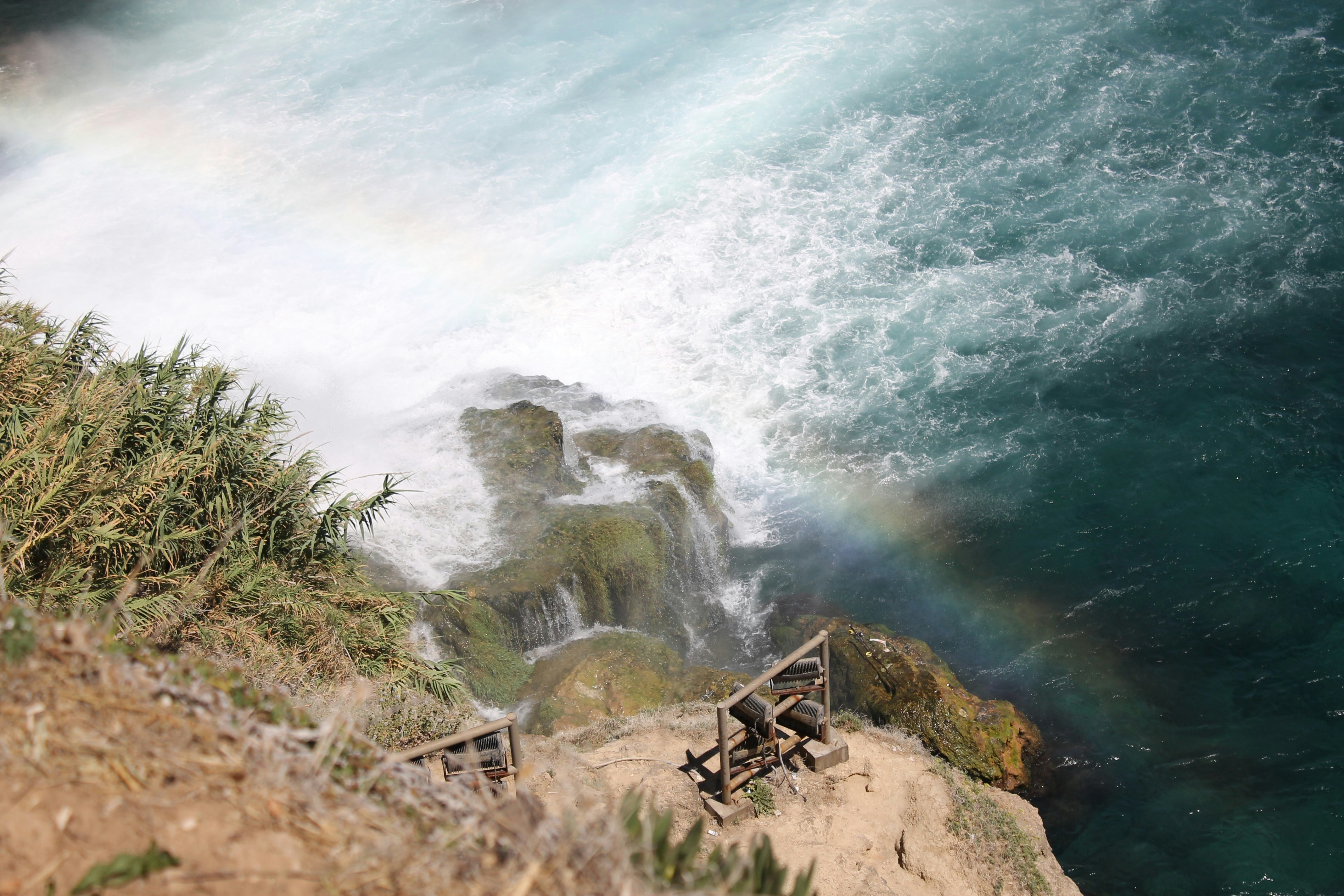 Rocky cliff path with stair railing descending toward turquoise surf and a subtle rainbow over the water.